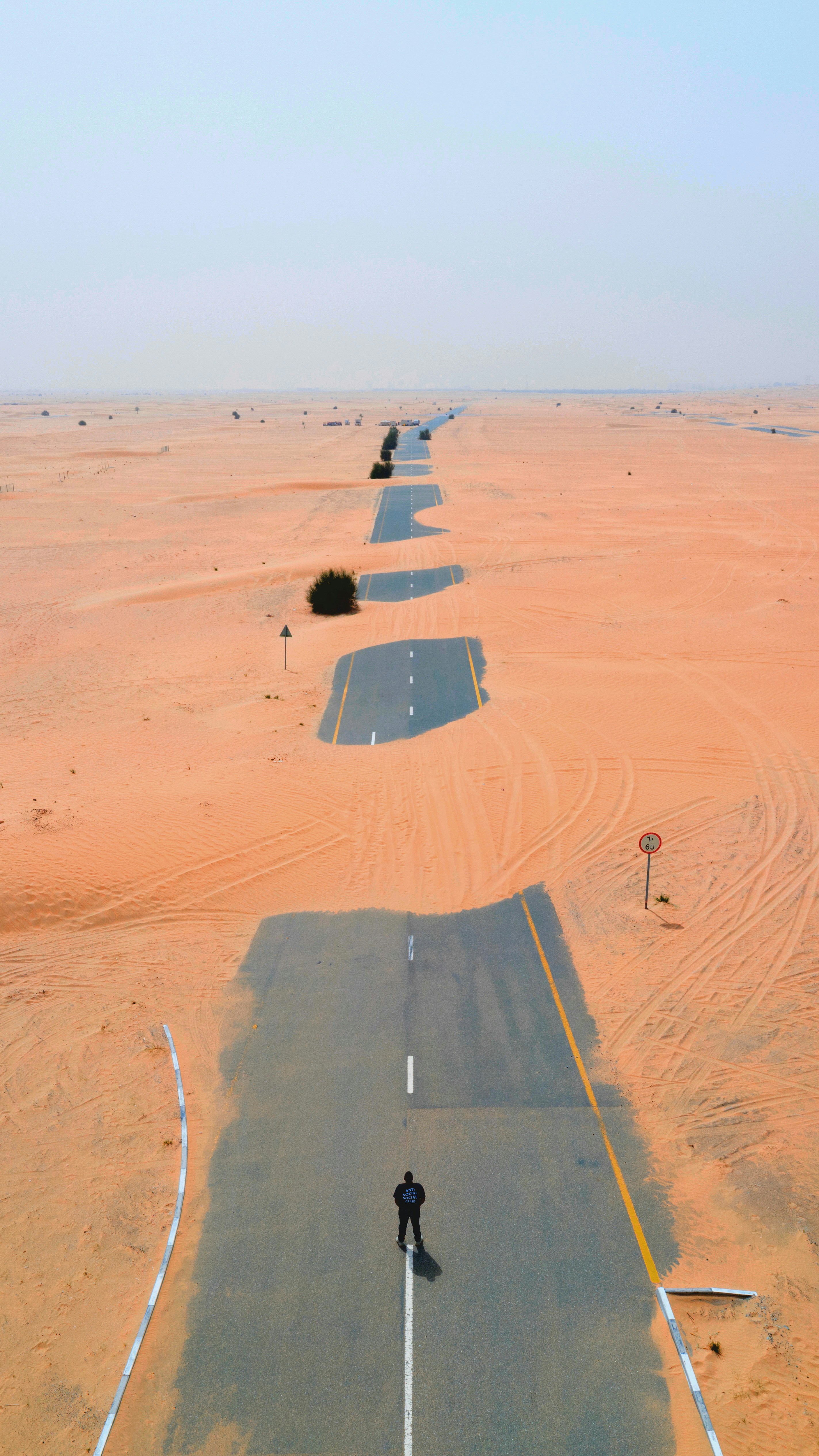 A lone figure stands on a deserted road, surrounded by vast stretches of sand dunes under a clear sky. The scene captures the stark contrast between the man-made road and the encroaching desert landscape.