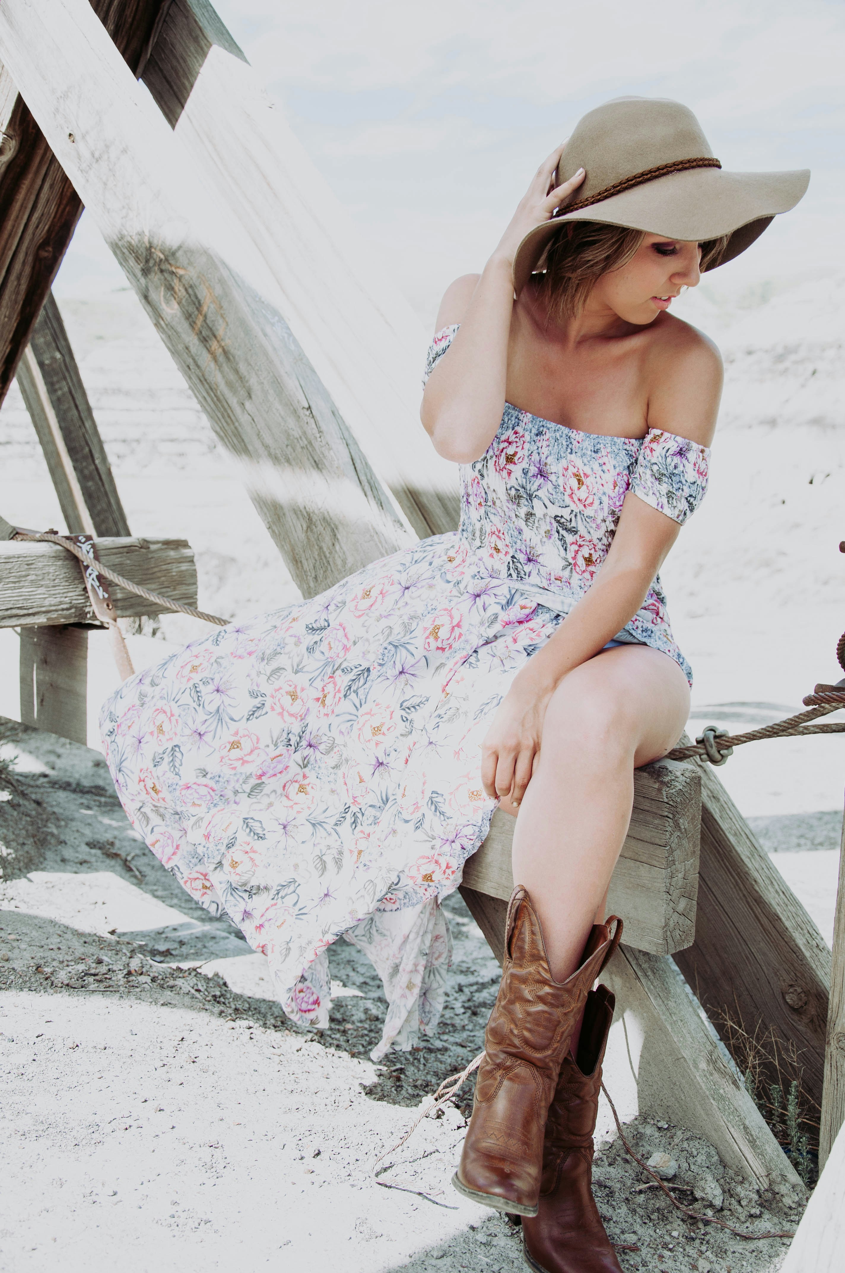 Woman in a floral dress and cowboy boots sits gracefully on a wooden structure, exuding a relaxed vibe amidst a natural landscape.