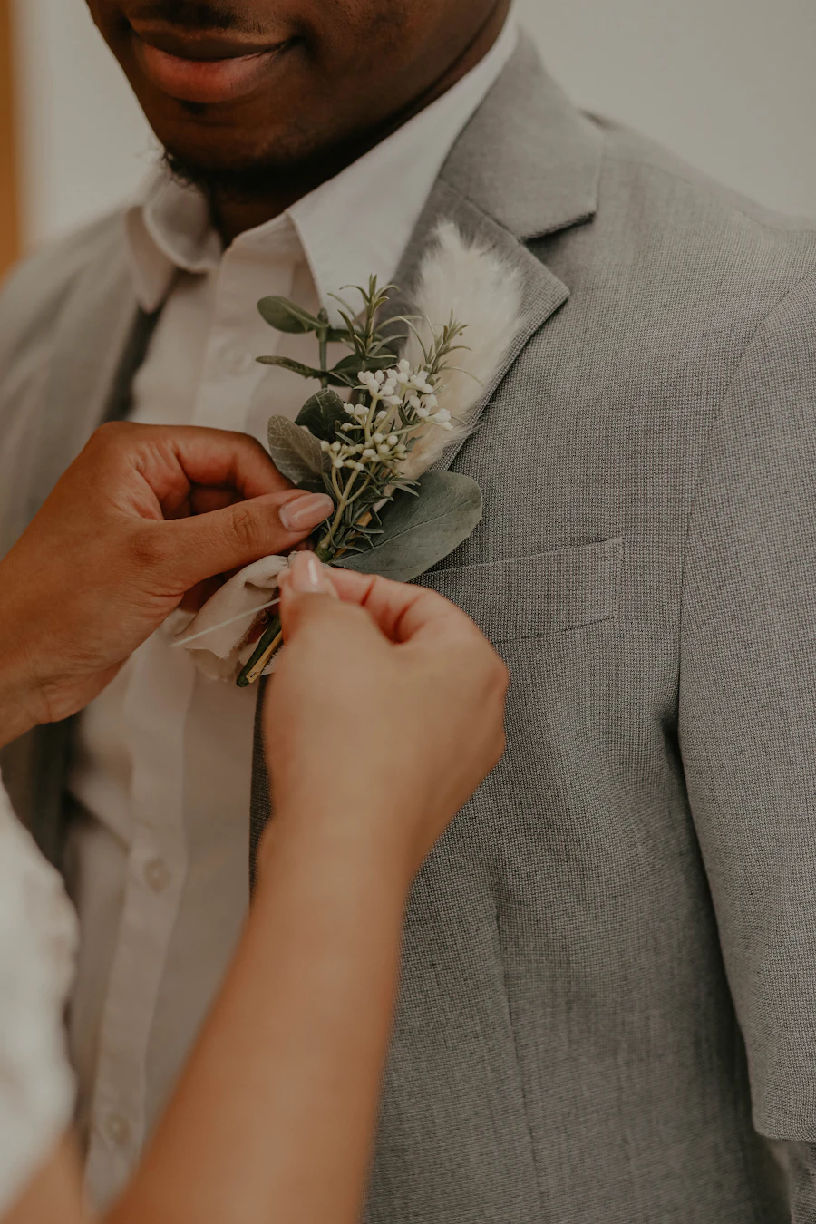Hands pin a small white boutonniere with greenery onto a man’s gray suit jacket. The cropped frame shows his lower face, white shirt, and the floral detail in soft warm light.