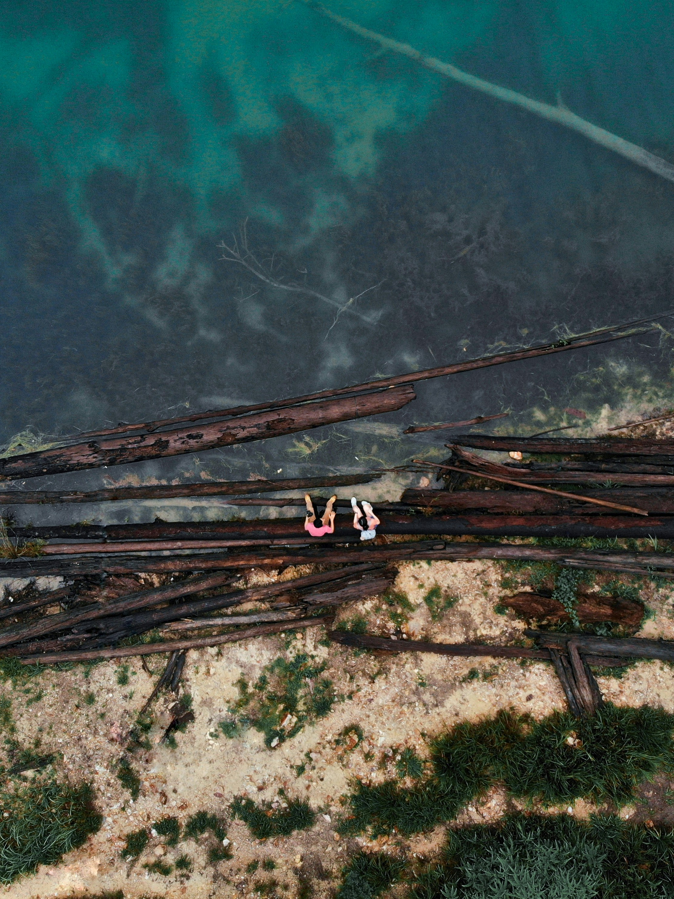 Two people lying on logs beside a turquoise lake, surrounded by greenery.