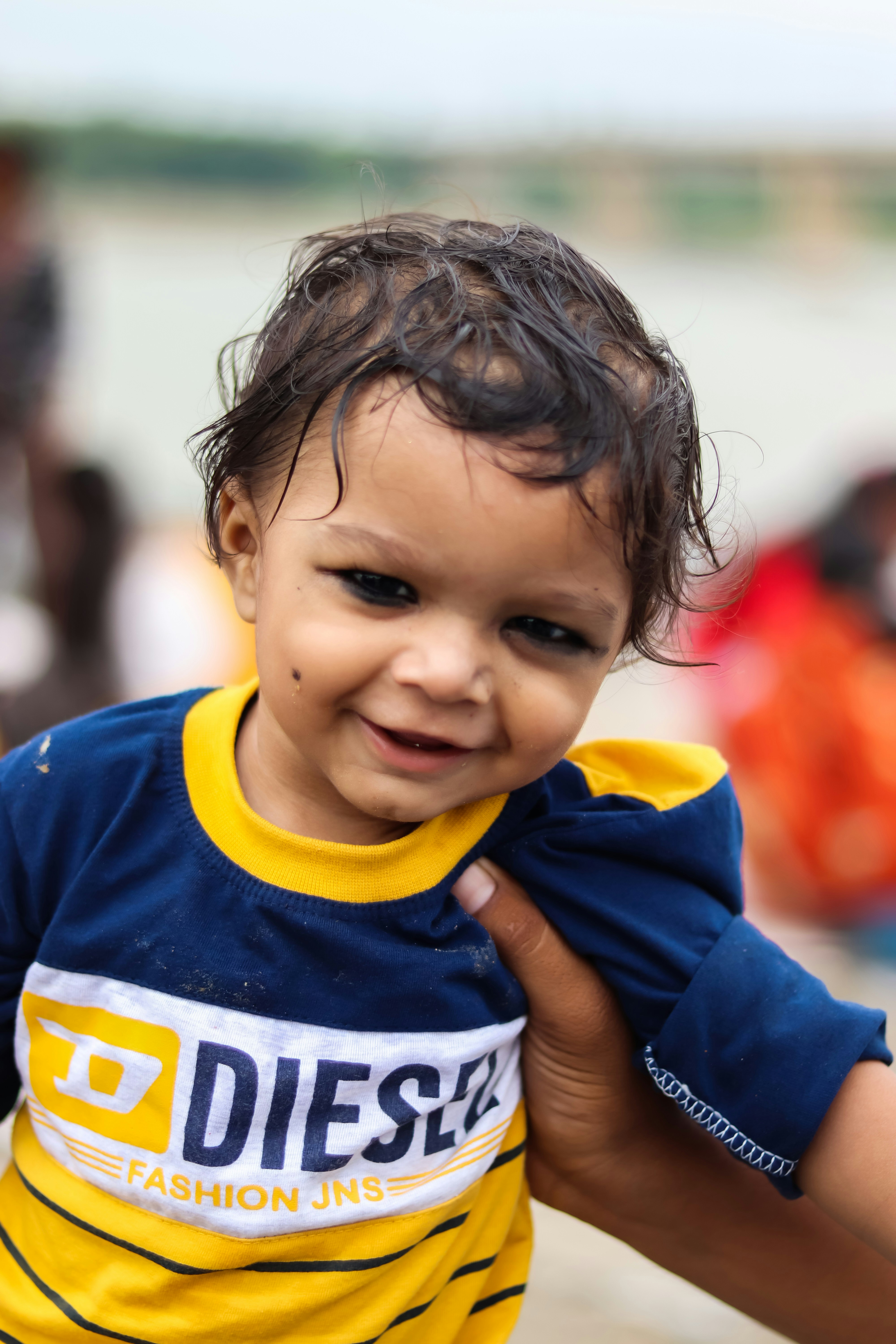 Smiling toddler in a blue-and-yellow Diesel shirt with a hand supporting from the side