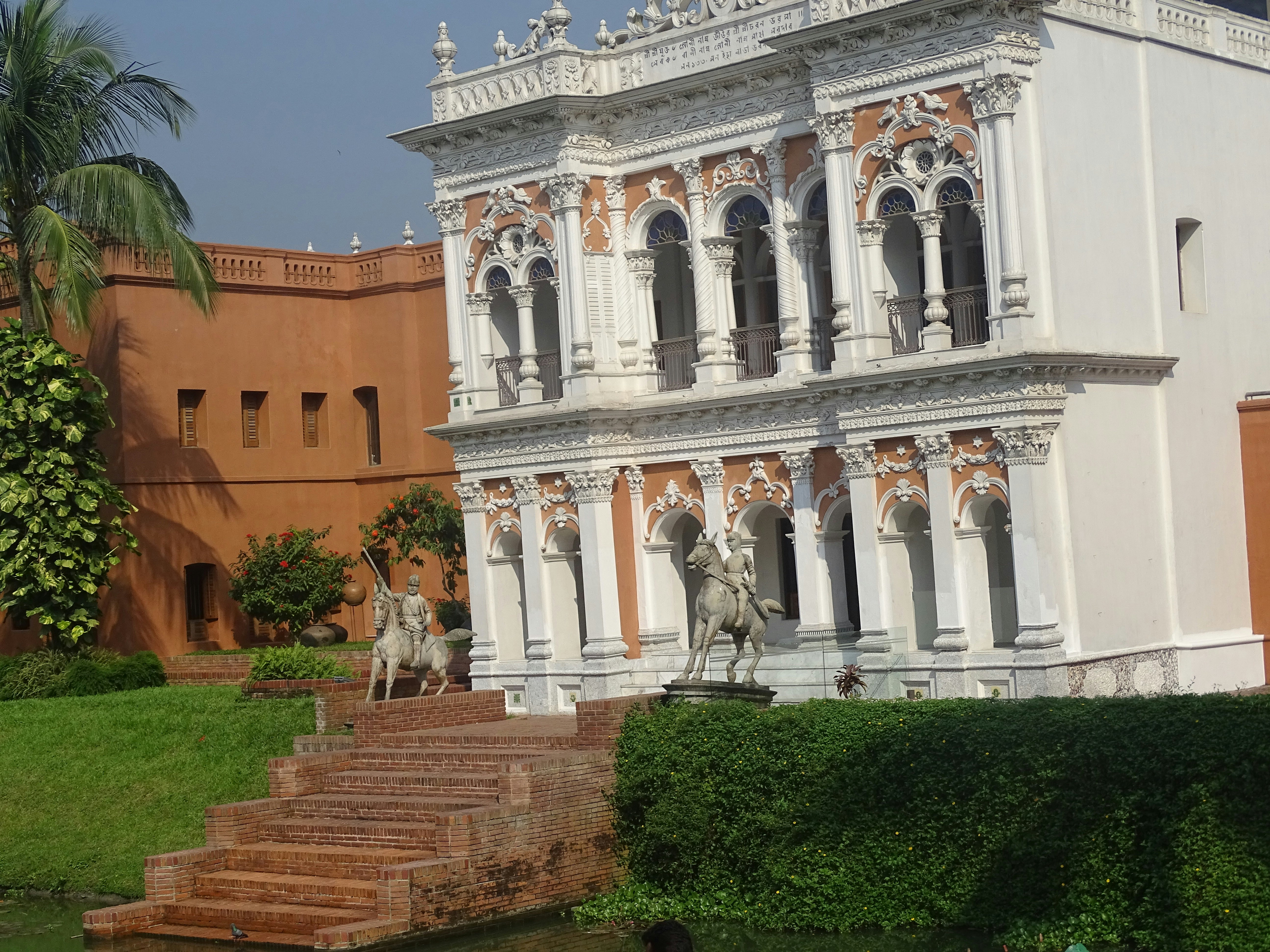 White neoclassical building with ornate columns beside a red brick structure surrounded by greenery.
