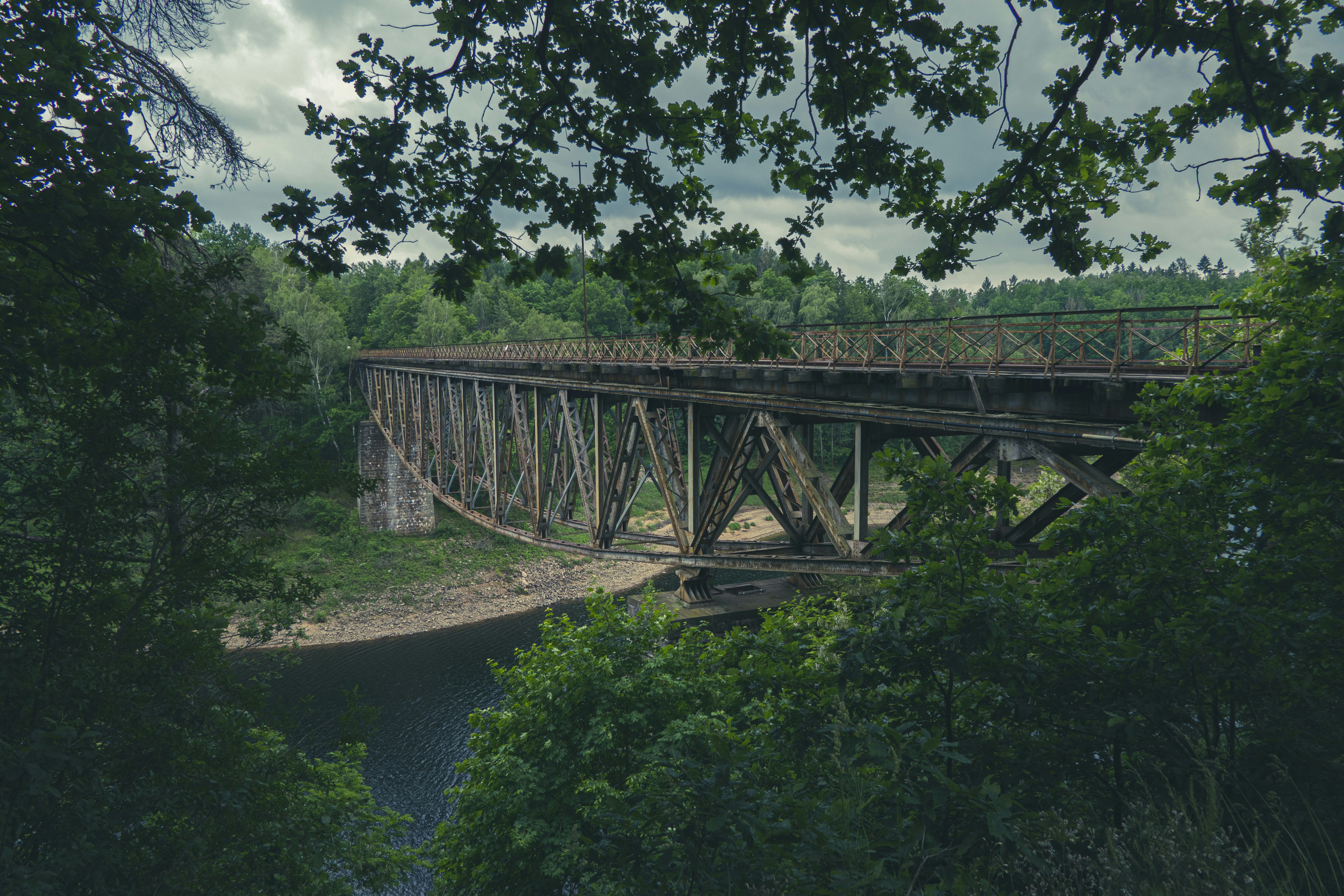 Rustic railway bridge spanning a tranquil river, framed by lush greenery and overcast skies.
