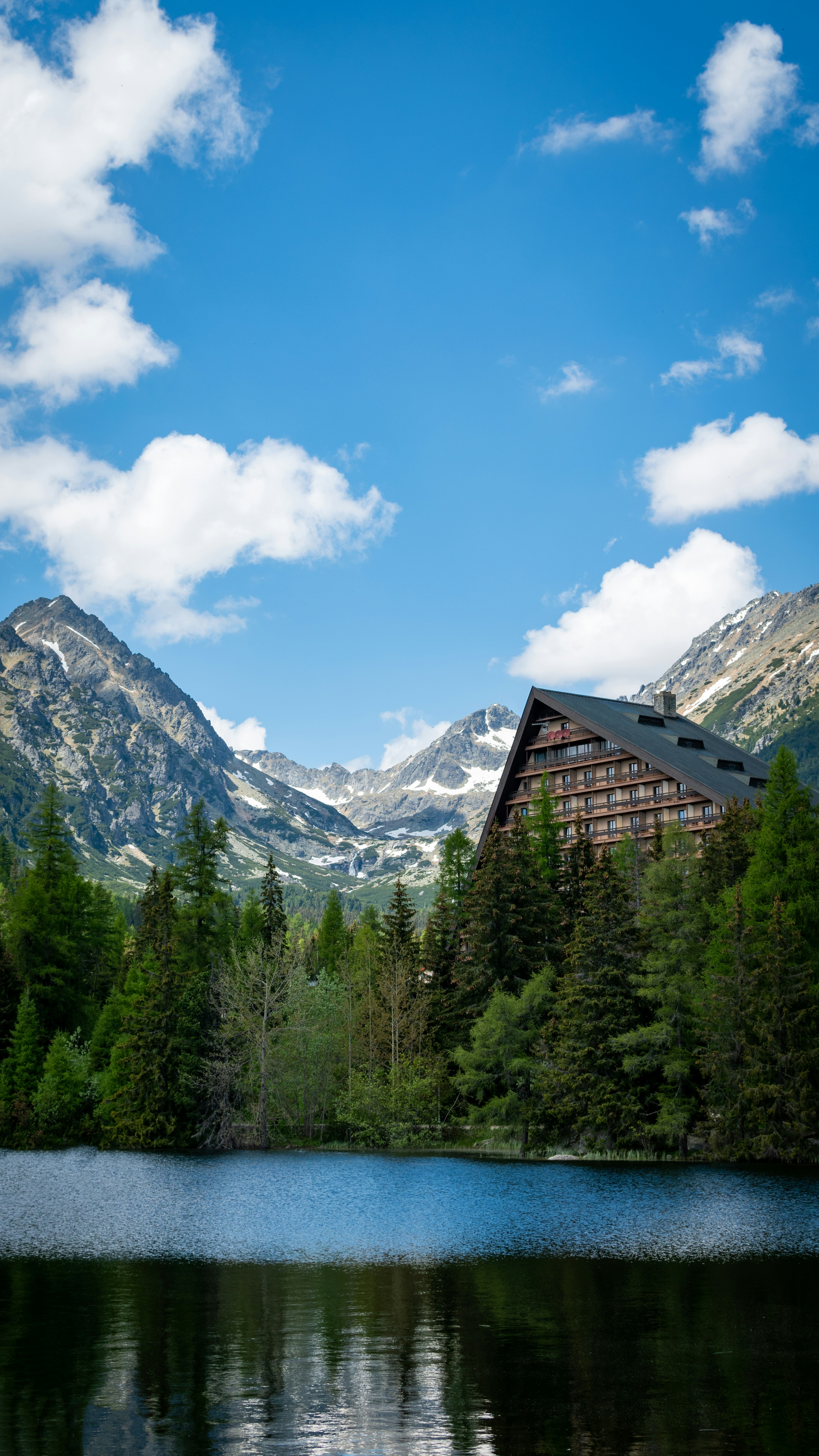 A modern lodge nestled among lush trees beside a tranquil lake, framed by majestic mountains and a vibrant blue sky.