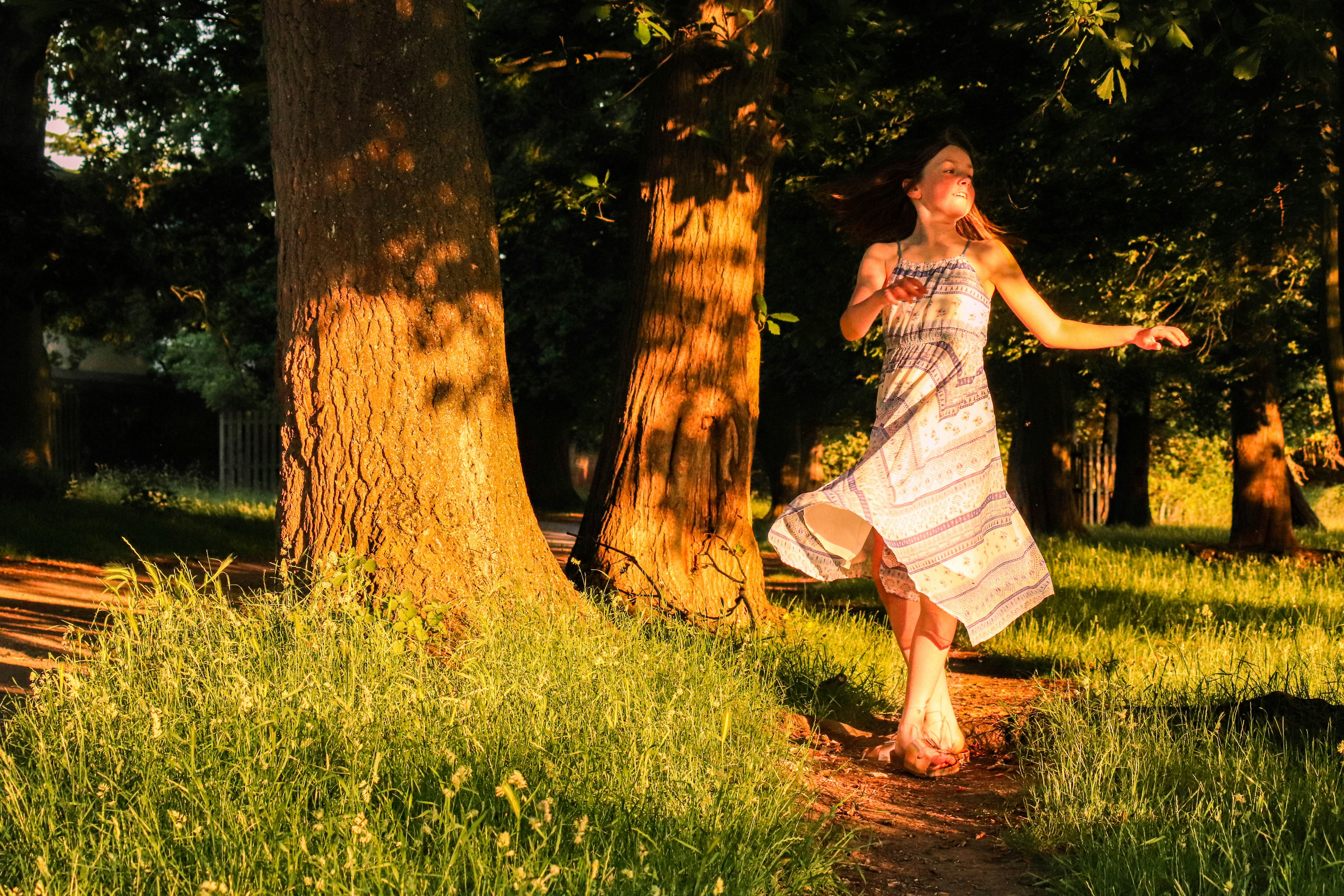 A woman twirls joyfully on a sunlit path surrounded by trees, with vibrant green grass beneath her feet.
