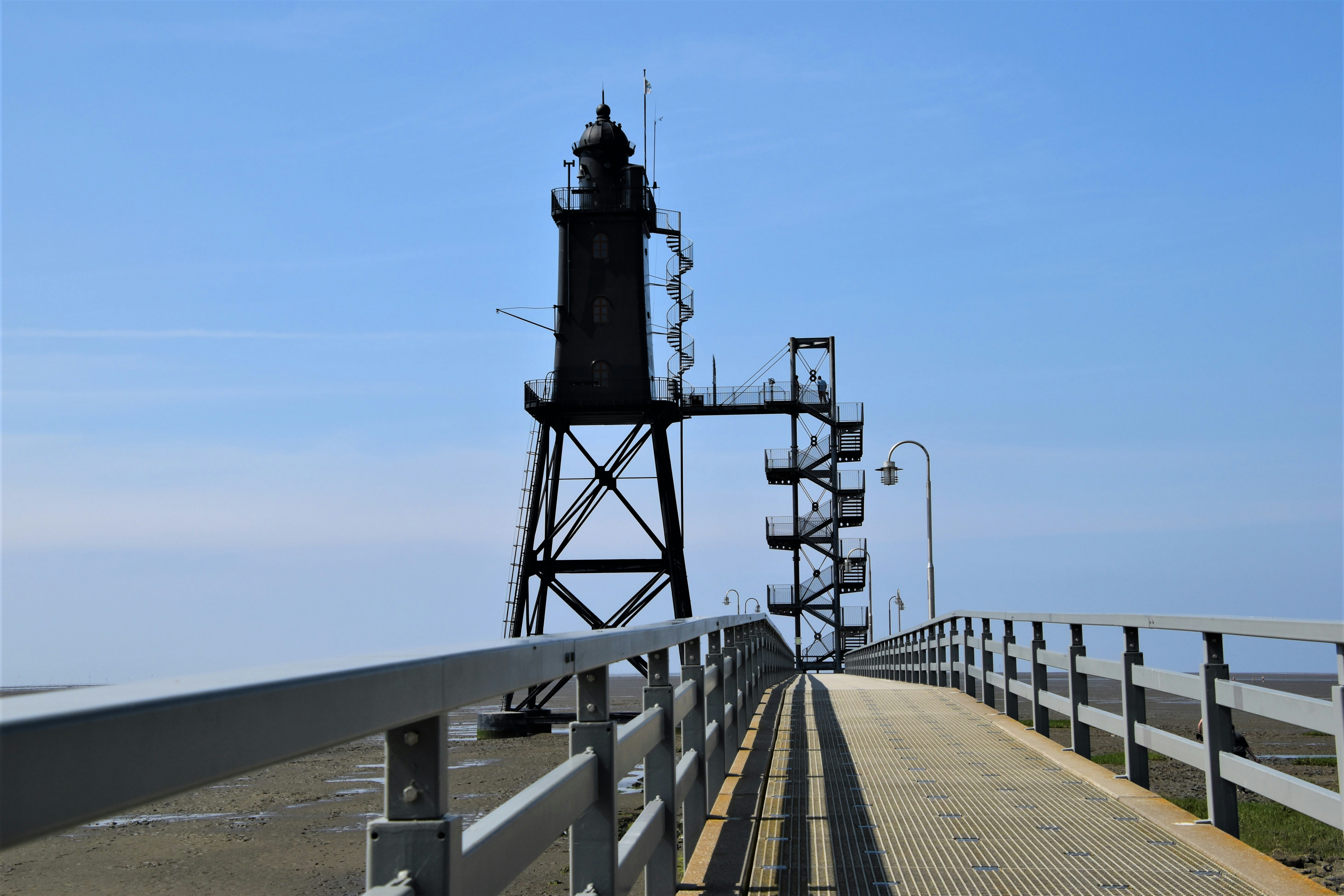 A vintage lighthouse stands tall beside a wooden walkway leading towards the shore, framed against a clear blue sky.