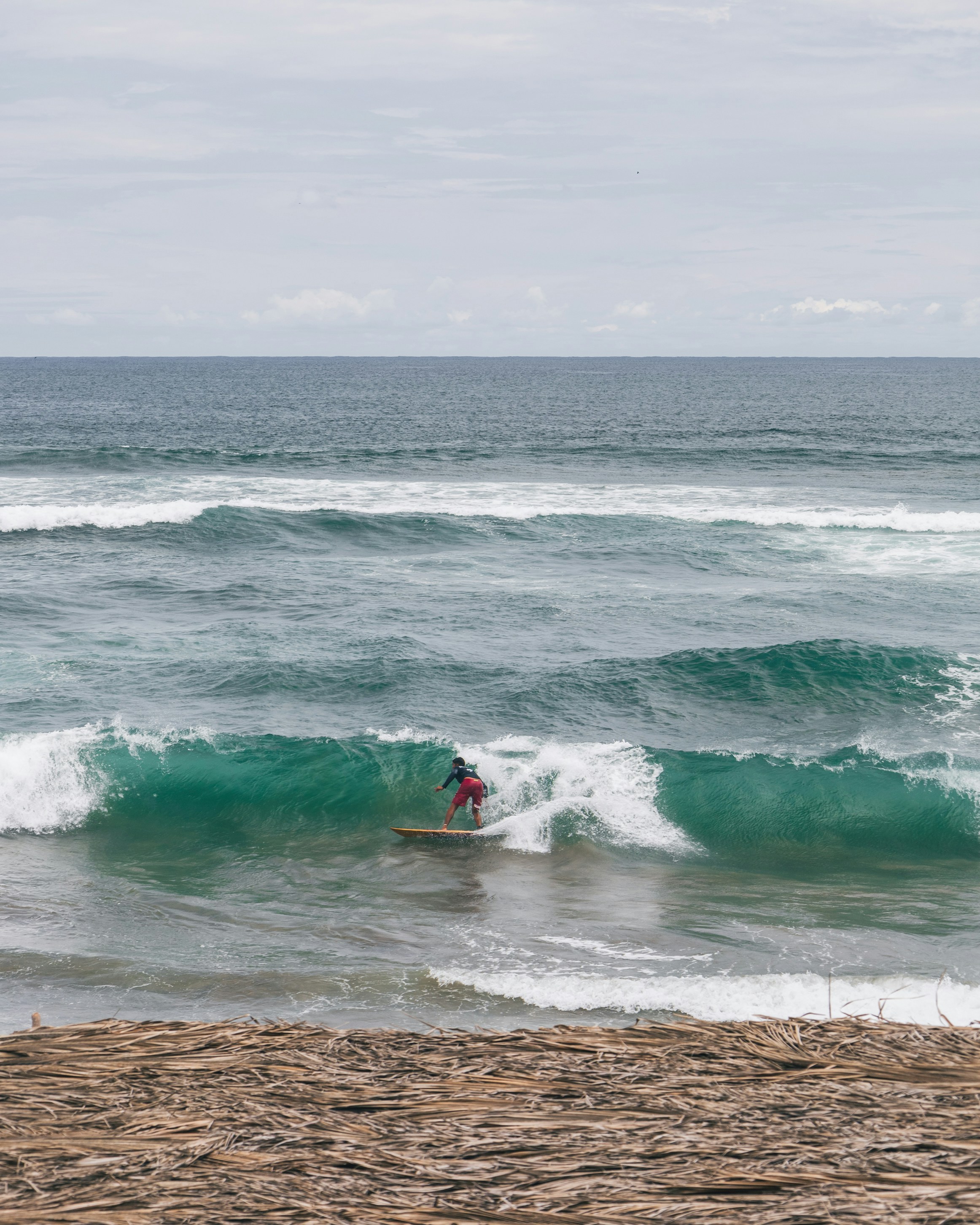 Surfer skillfully navigating a vibrant wave along a sandy beach, with a backdrop of rolling ocean waves and a cloudy sky.