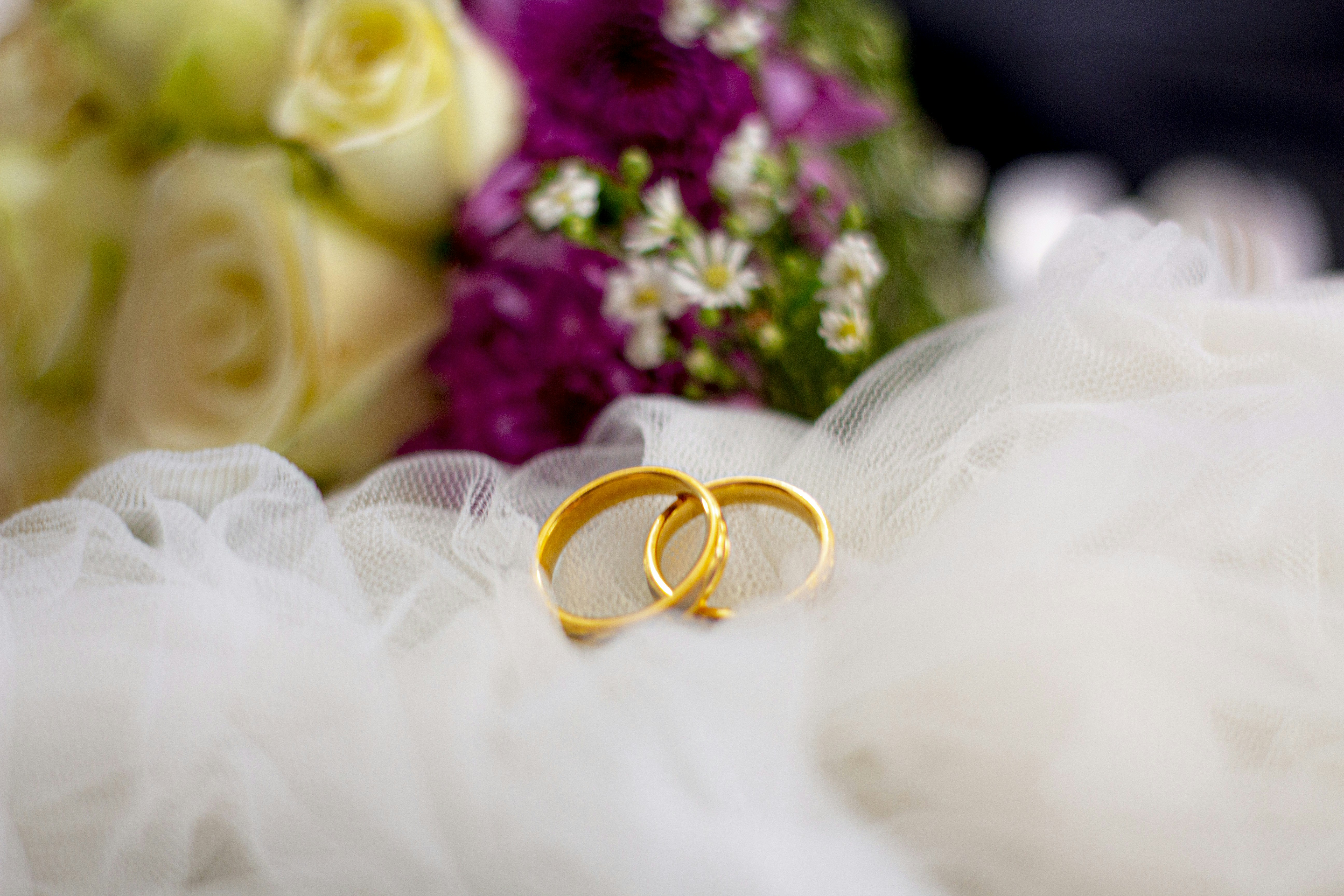 Two intertwined gold wedding rings resting on delicate tulle, surrounded by a bouquet of roses and vibrant flowers.