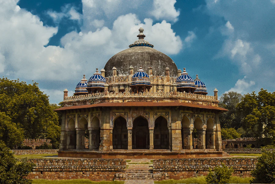 An old domed stone mausoleum with blue-tiled turrets stands centered in a landscaped garden beneath a cloudy sky. Trees and low stone walls frame the structure.