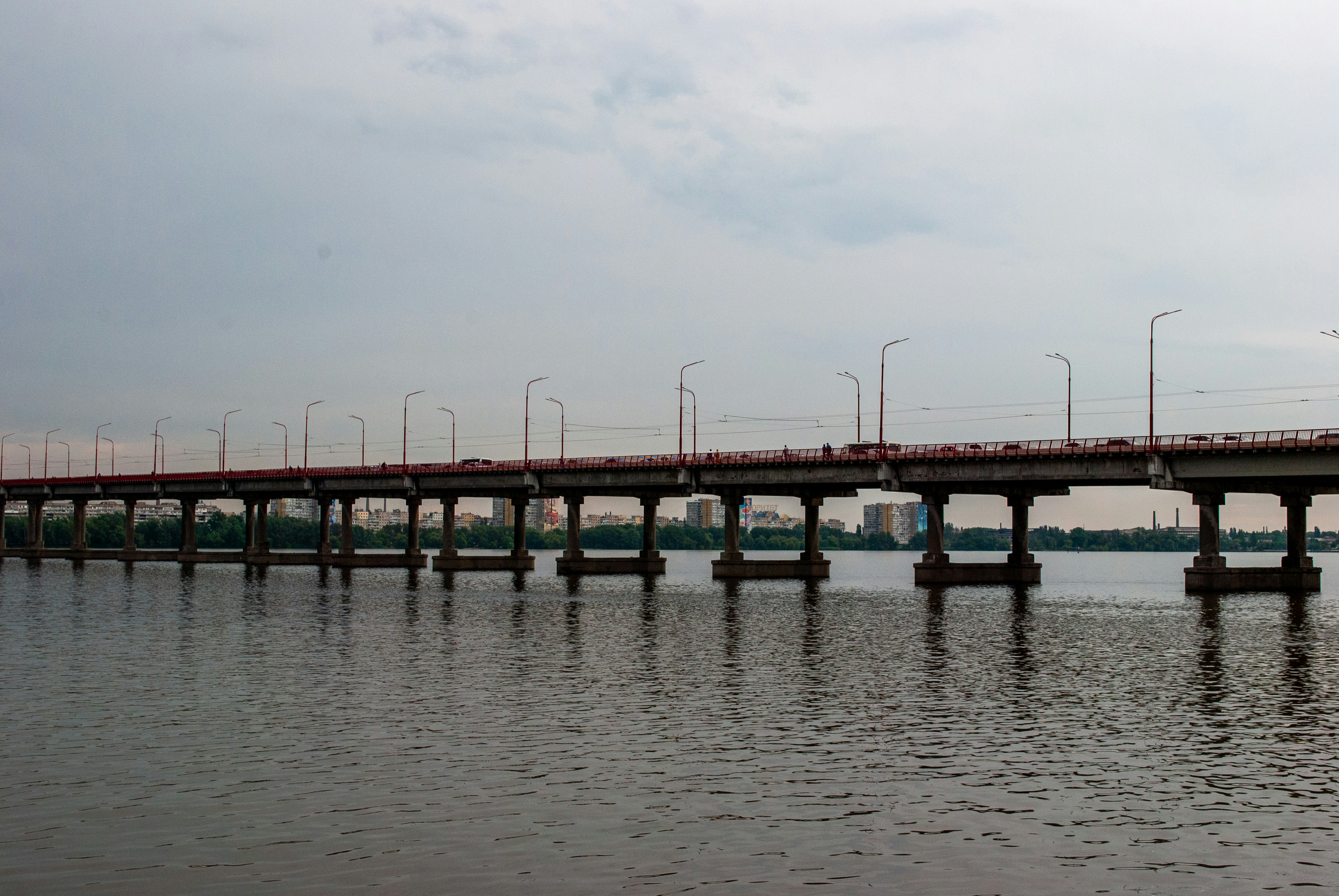 A long bridge spans a calm river, with reflections shimmering on the water's surface. The scene captures the intersection of urban life and nature.