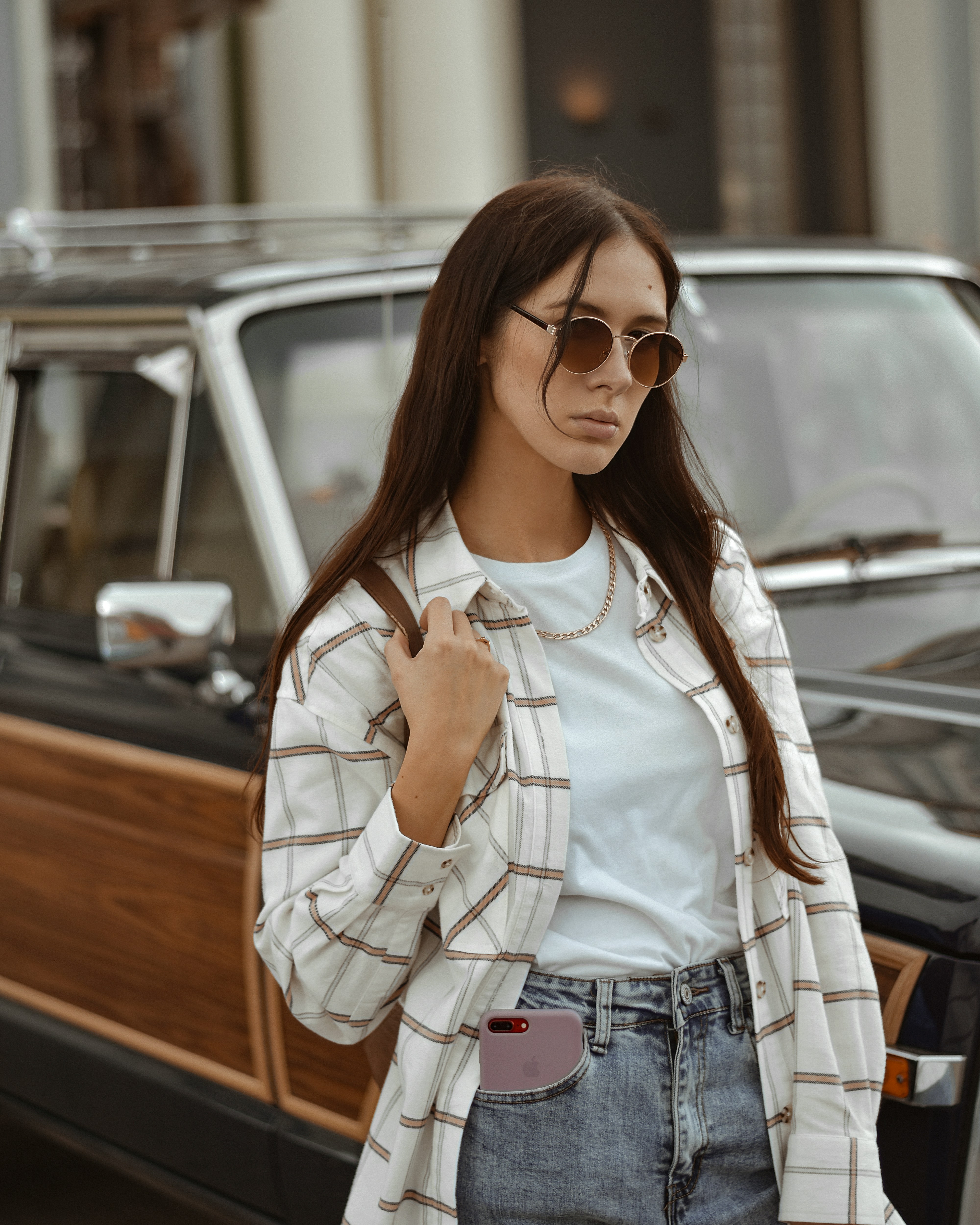 Fashionable woman in plaid shirt and sunglasses stands beside a vintage car, exuding a contemporary urban vibe.