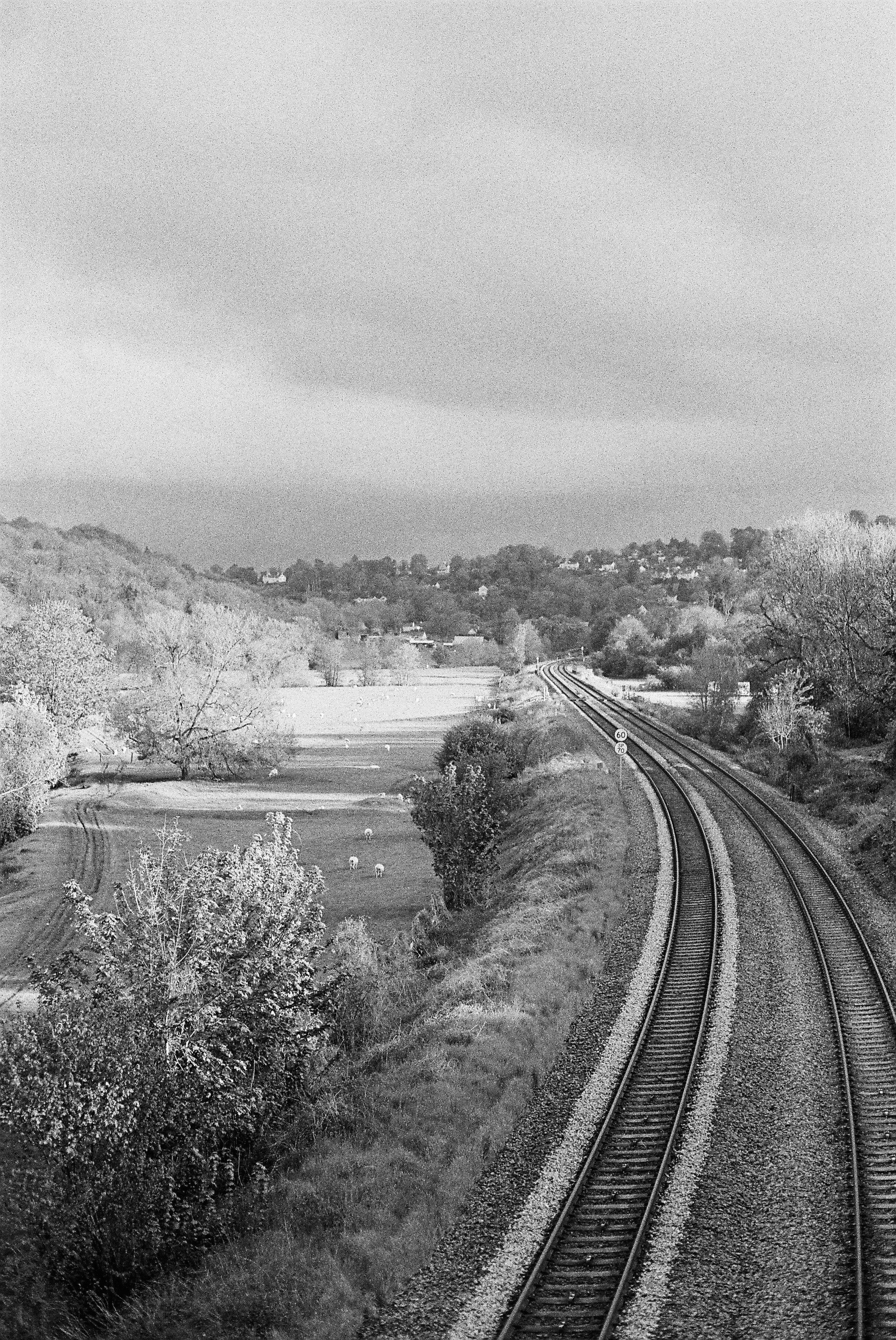 Monochrome photograph of a railway curve winding through a tranquil countryside with trees and distant hills.