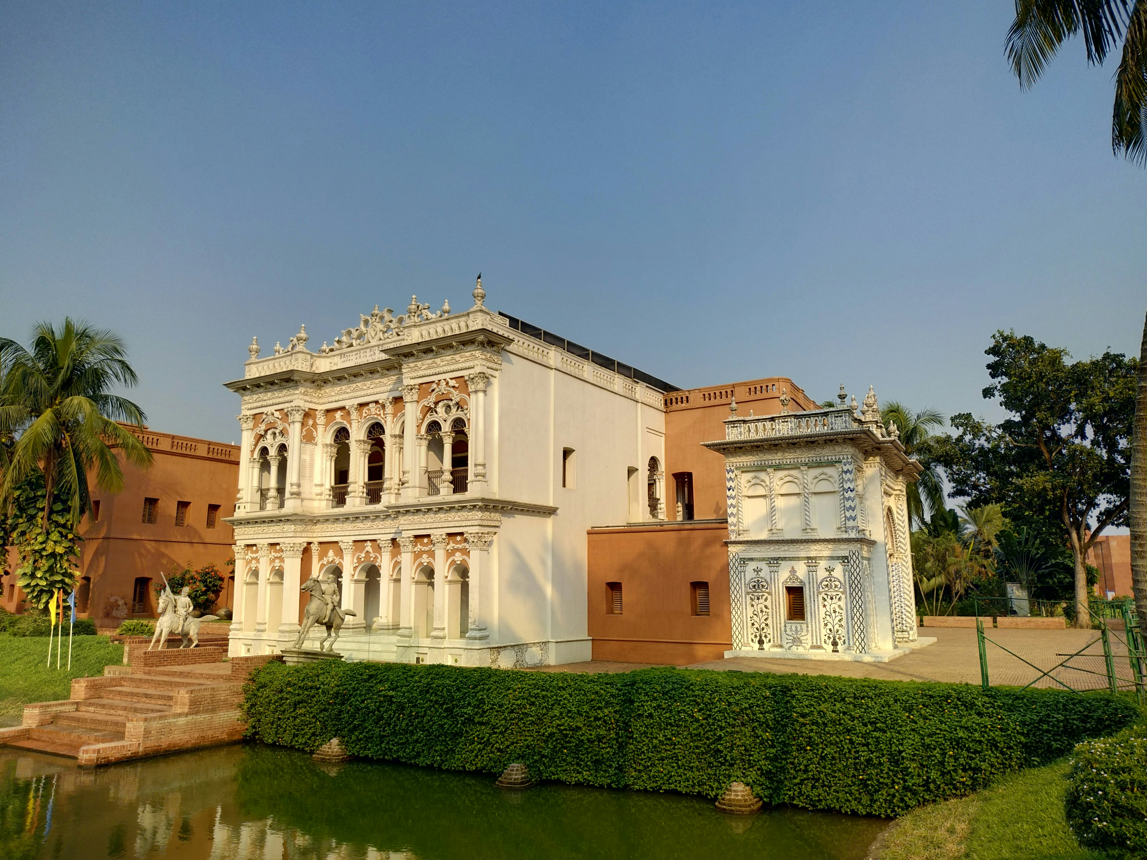 Historic white mansion with ornate details beside a serene pond under a clear blue sky.