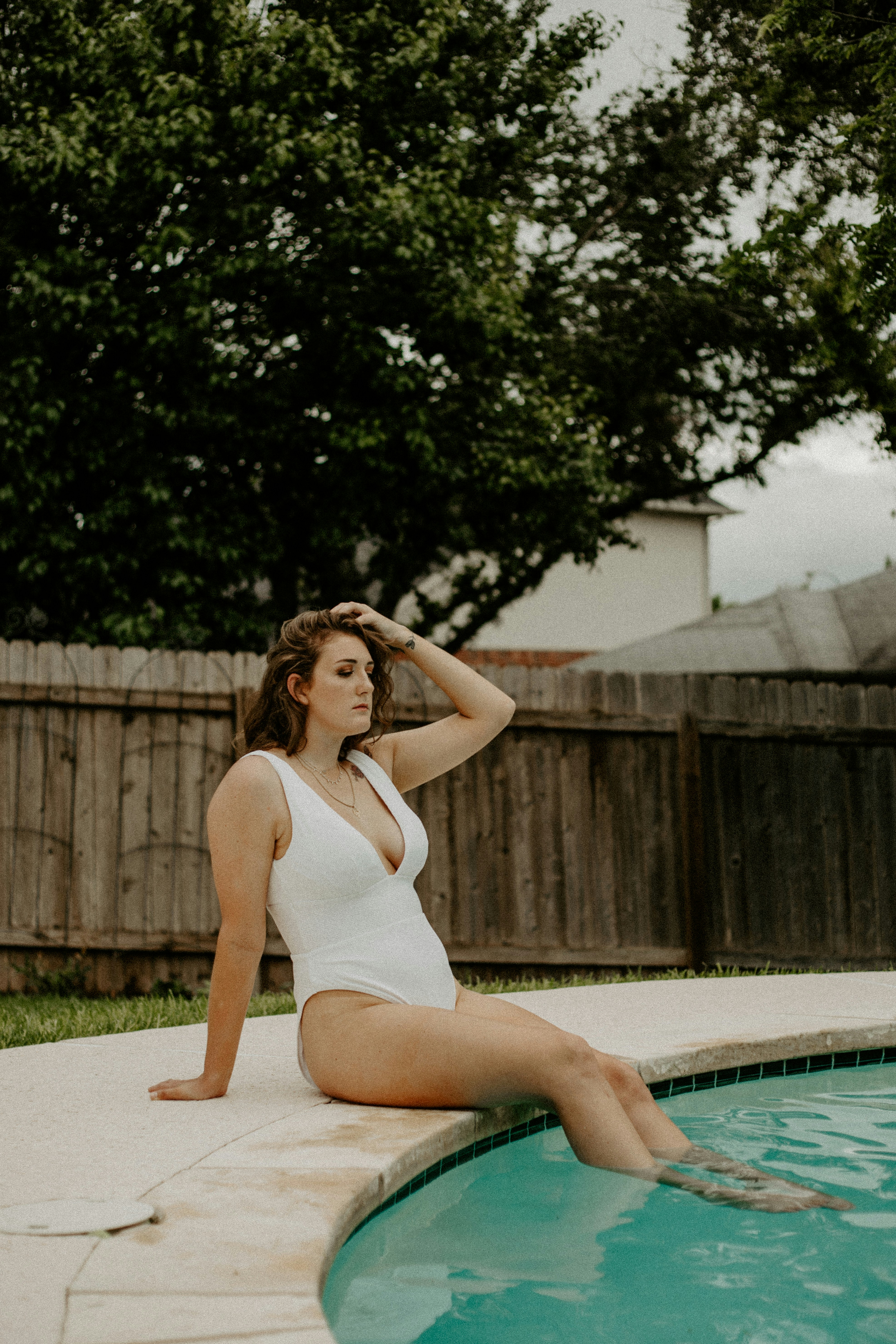 A woman in a white swimsuit relaxes by a pool, her hand resting on her head, surrounded by lush greenery and a wooden fence.
