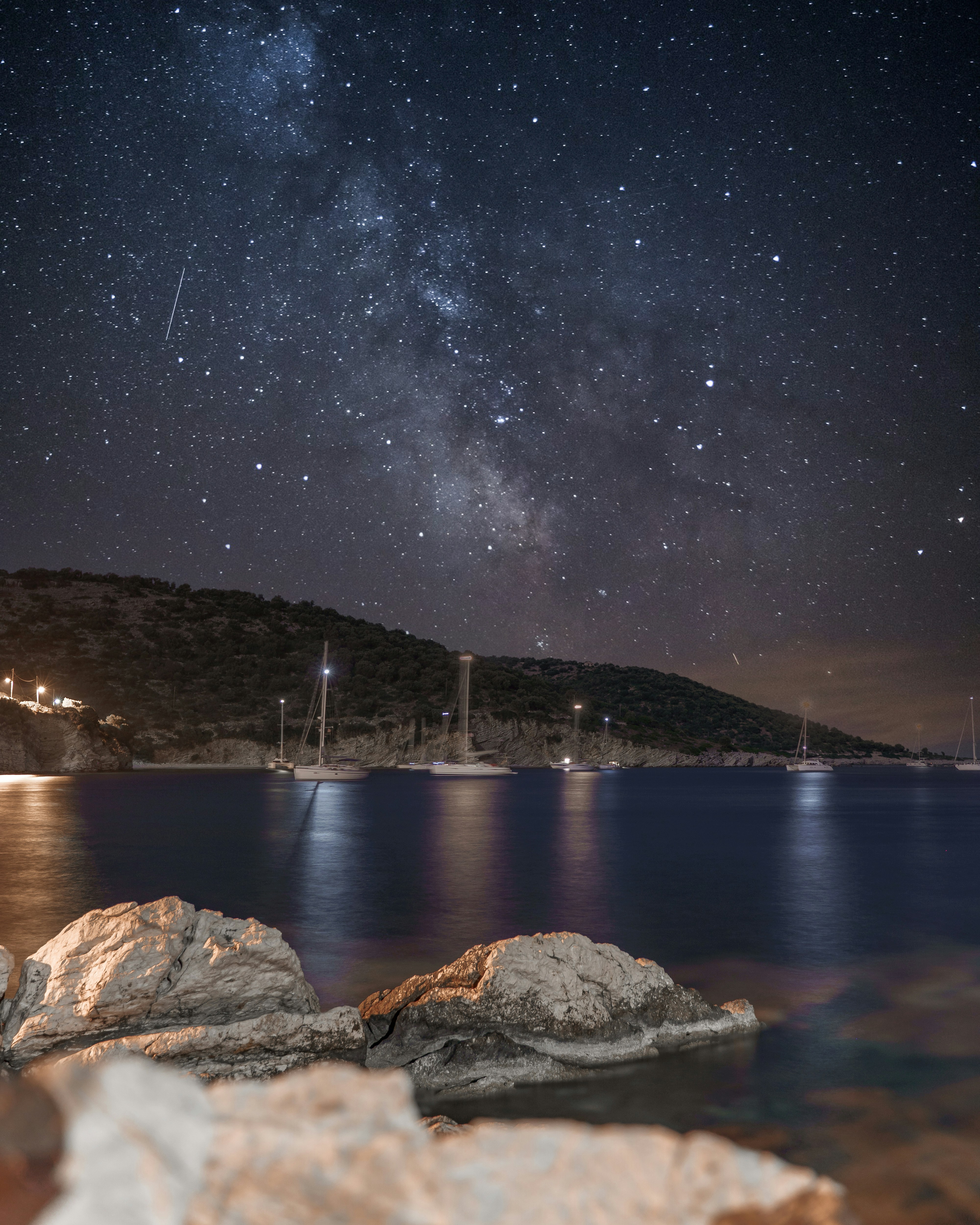 A serene coastal scene under a starry sky, featuring the Milky Way and illuminated boats anchored in calm waters. The rocky shoreline adds depth to the composition.