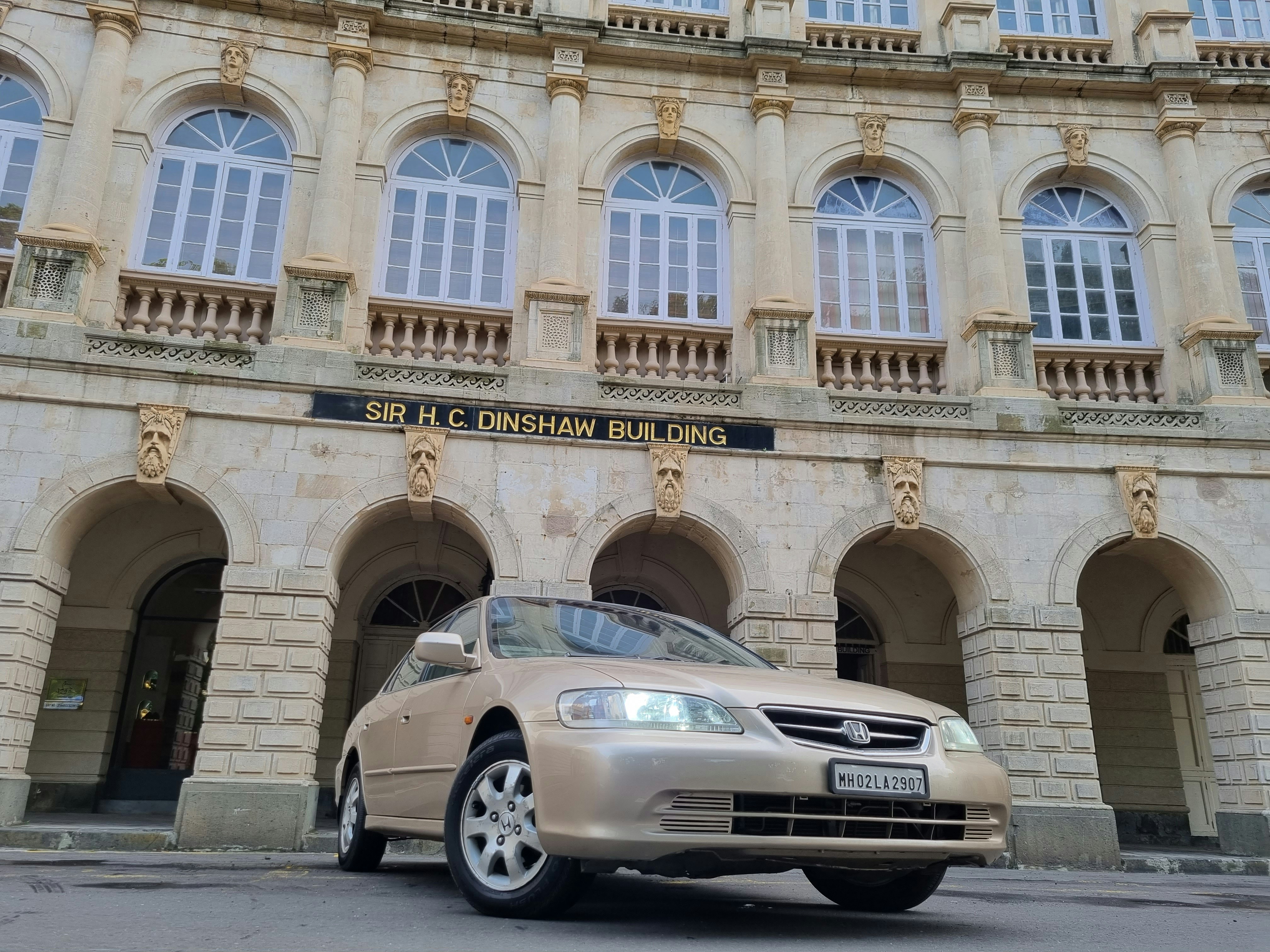 A classic beige sedan parked in front of an ornate historic building with arched windows and columns during the day.