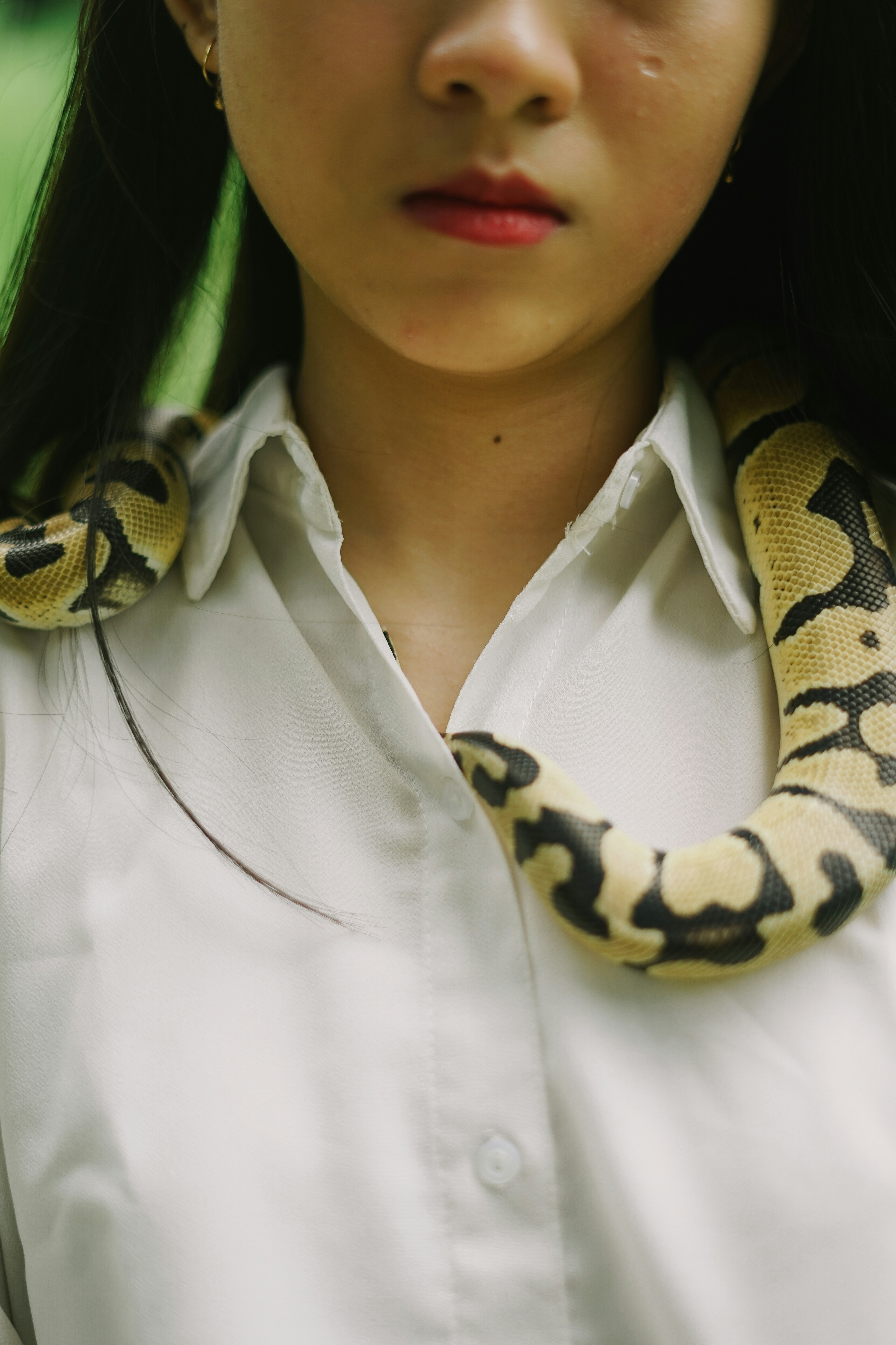Close-up portrait of a woman with a leopard-pattern snake draped around a white shirt, set against a soft green bokeh background.