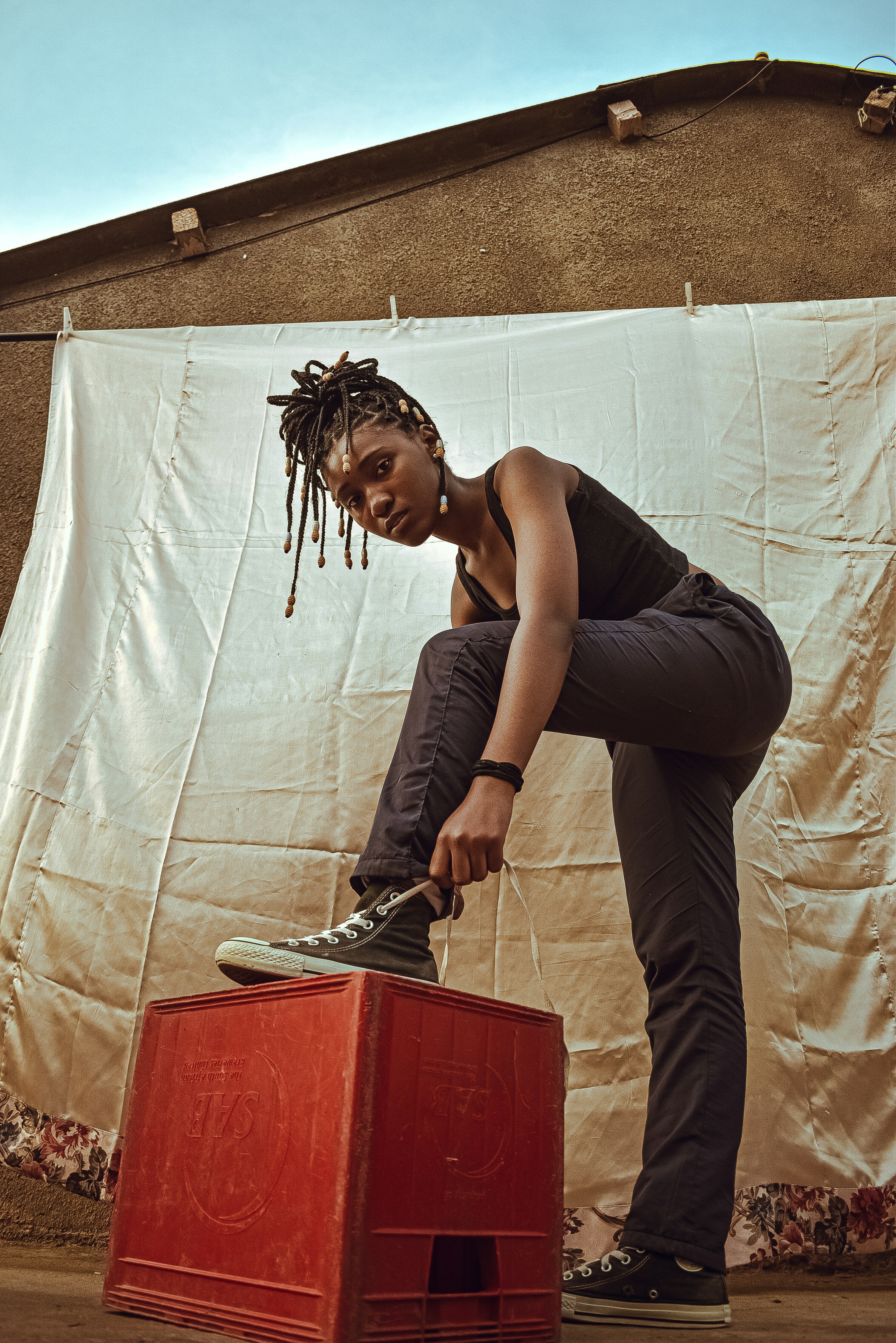 Young woman adjusting her shoe while perched on a red crate, set against a backdrop of a white fabric. The scene captures a blend of casual style and dynamic movement.