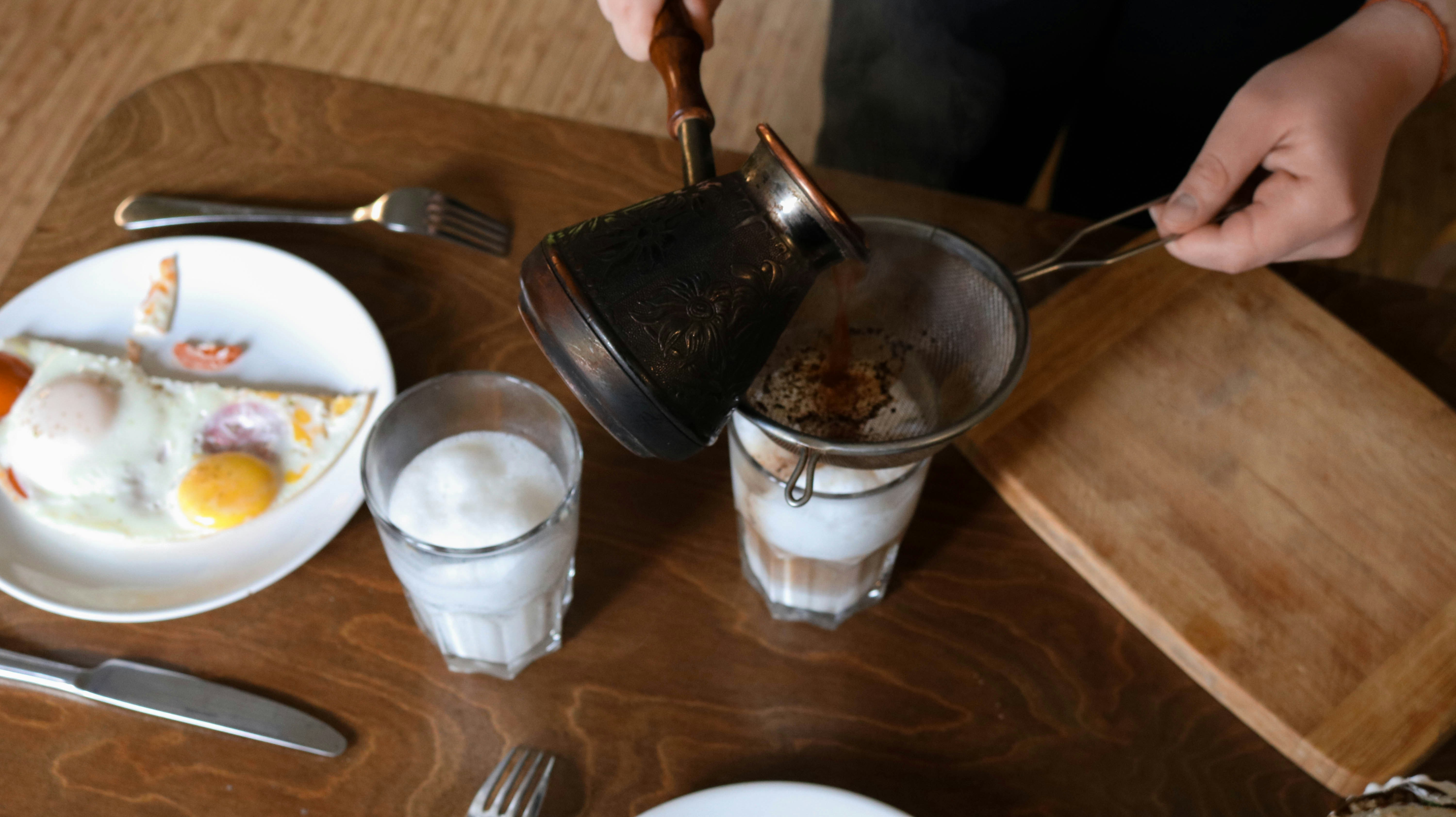 Traditional coffee being poured through a filter into a glass, accompanied by a plate of eggs and toast. The setting evokes a cozy breakfast atmosphere.