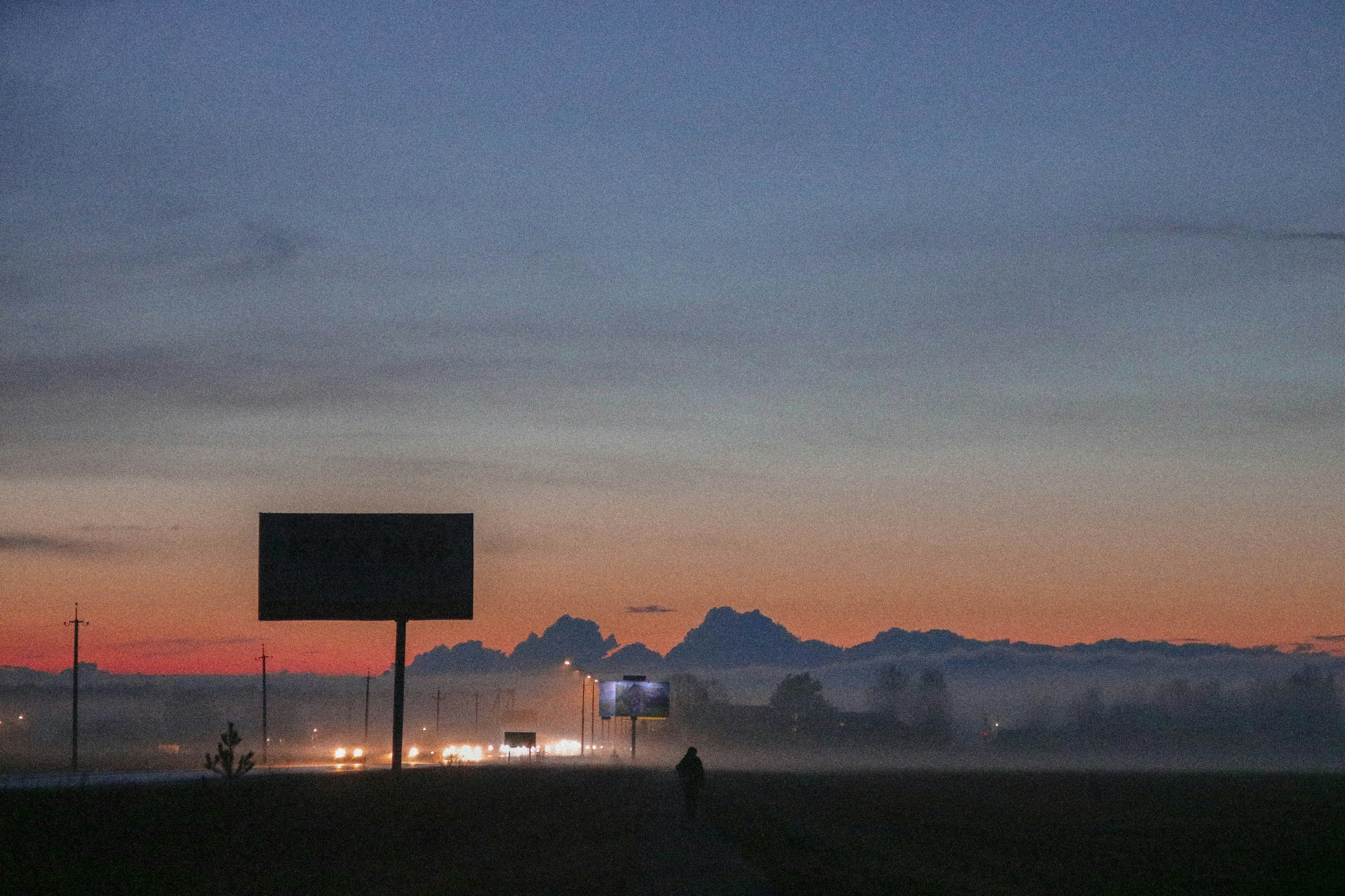 A solitary figure walks along a foggy road at twilight, silhouetted against distant mountains and glowing billboards. The scene captures the serene transition from day to night.