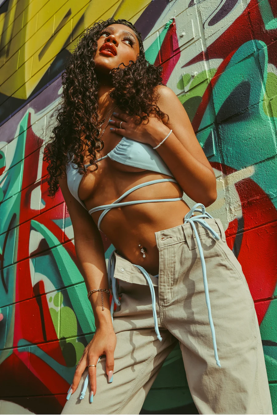 A woman poses in front of a vivid graffiti wall, looking upward with one hand at her chest. She wears a light blue strappy top and beige pants under bright sunlight.