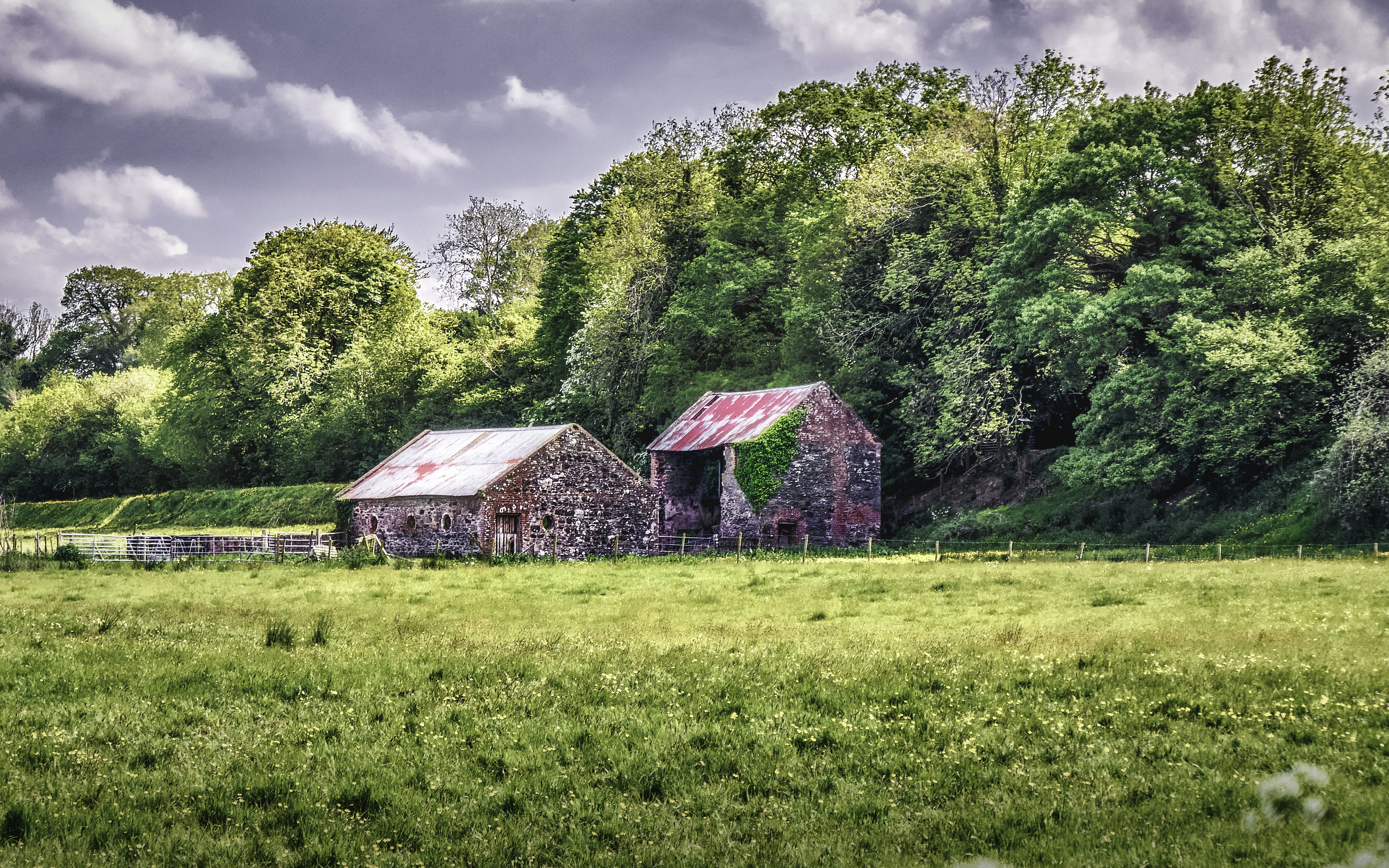 Abandoned stone barn and shed surrounded by lush greenery under a cloudy sky. The structures exhibit signs of age, blending with the serene landscape.
