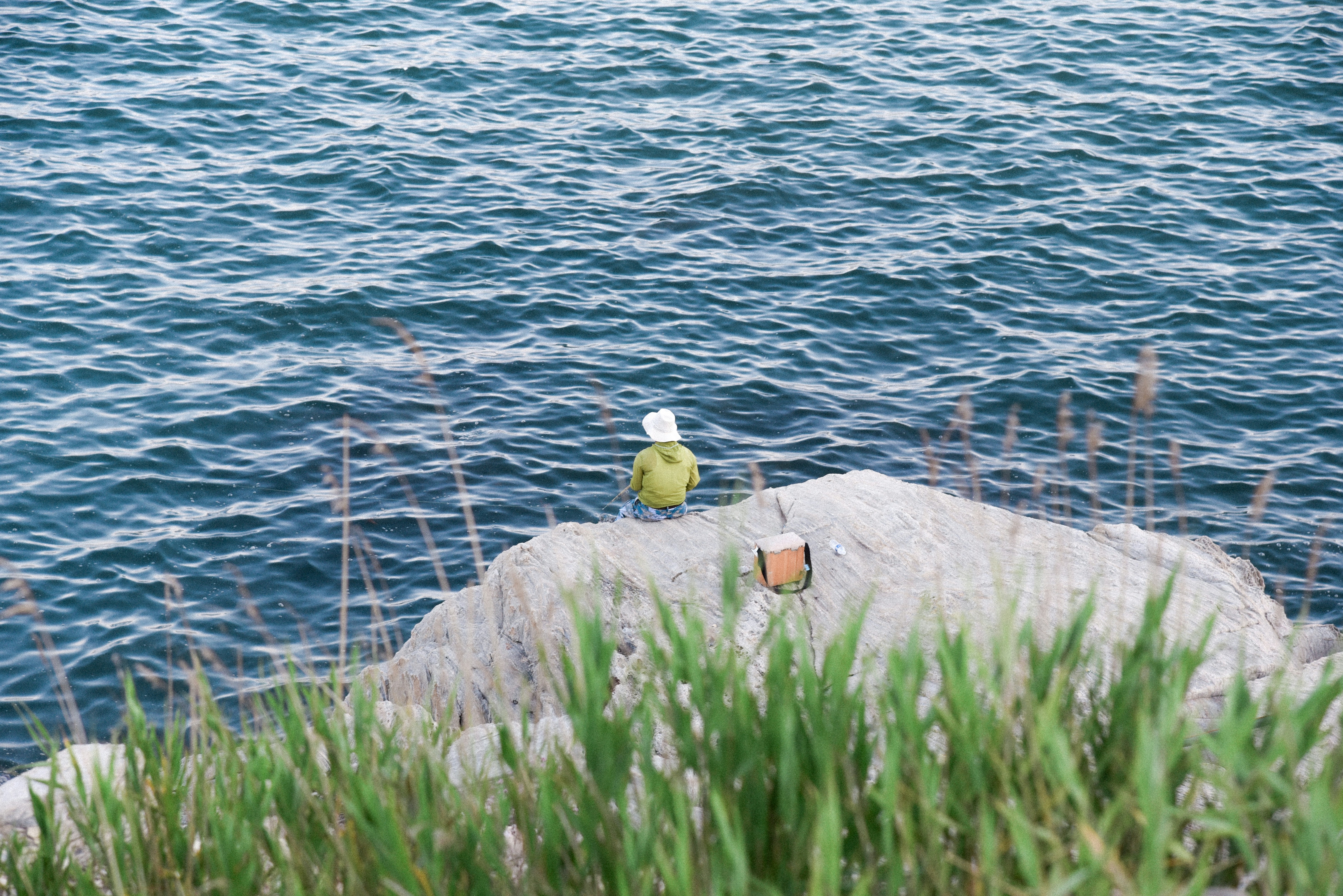 Person sitting on a rock by the water, surrounded by tall grass, gazing at the waves. A fishing bucket is placed nearby.