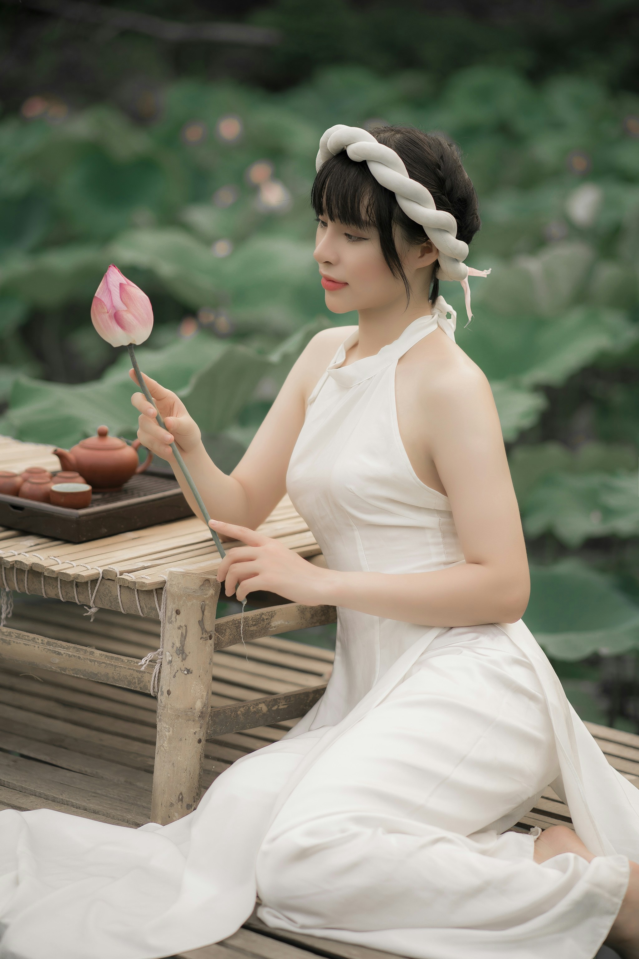 A woman in a flowing white dress delicately holds a pink lotus flower, surrounded by lush green foliage and a tea set. The scene evokes tranquility and harmony with nature.