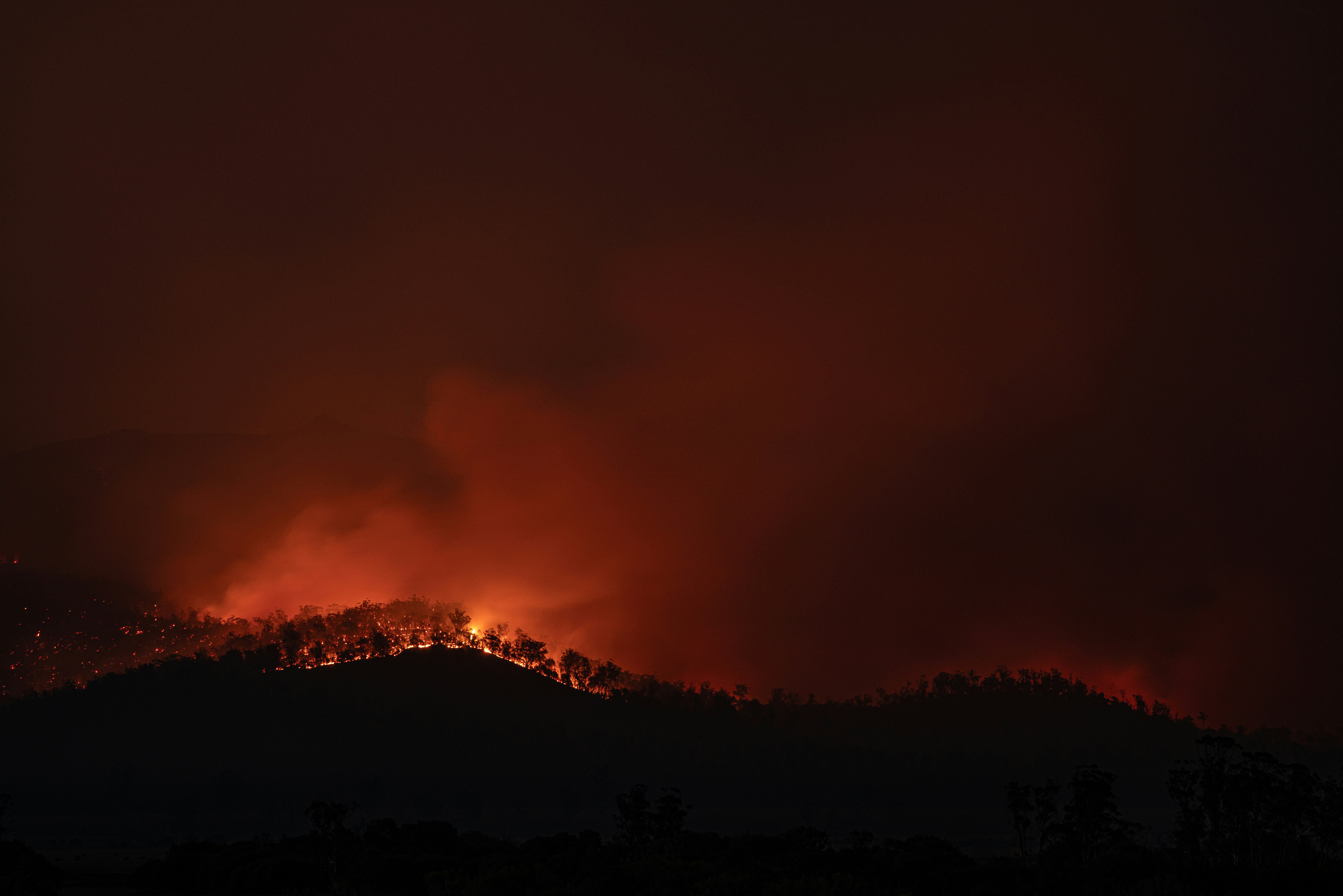 This dramatic image captures a forest fire raging across a hillside, with fiery hues illuminating the night sky. The composition highlights the stark contrast between the bright orange flames and the dark silhouettes of trees, creating a scene of both beauty and devastation. The rich color palette and low-light conditions enhance the intensity, drawing attention to the fire's raw power and atmospheric glow.