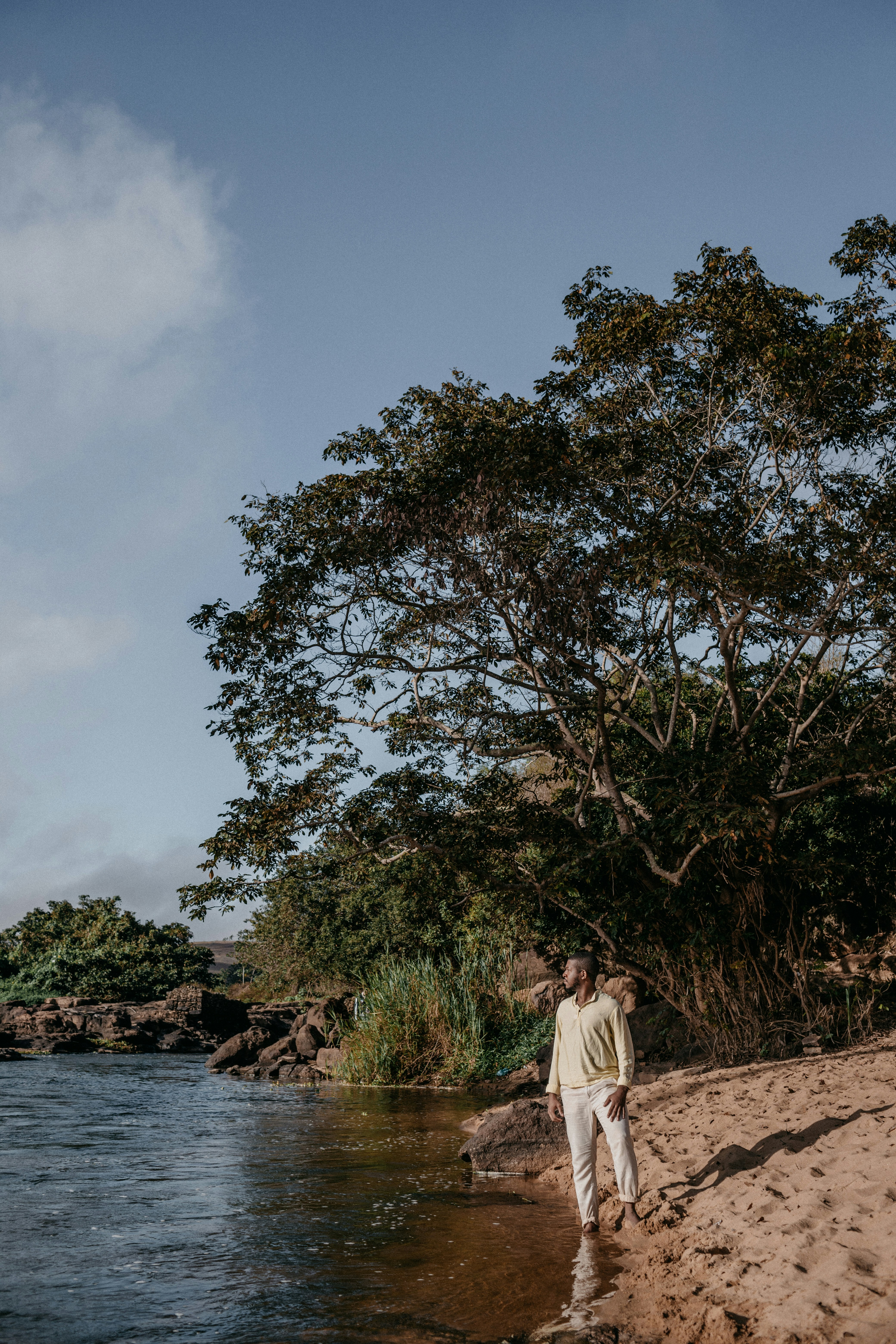 A person stands thoughtfully by the water's edge, surrounded by lush greenery and a tranquil riverbank.