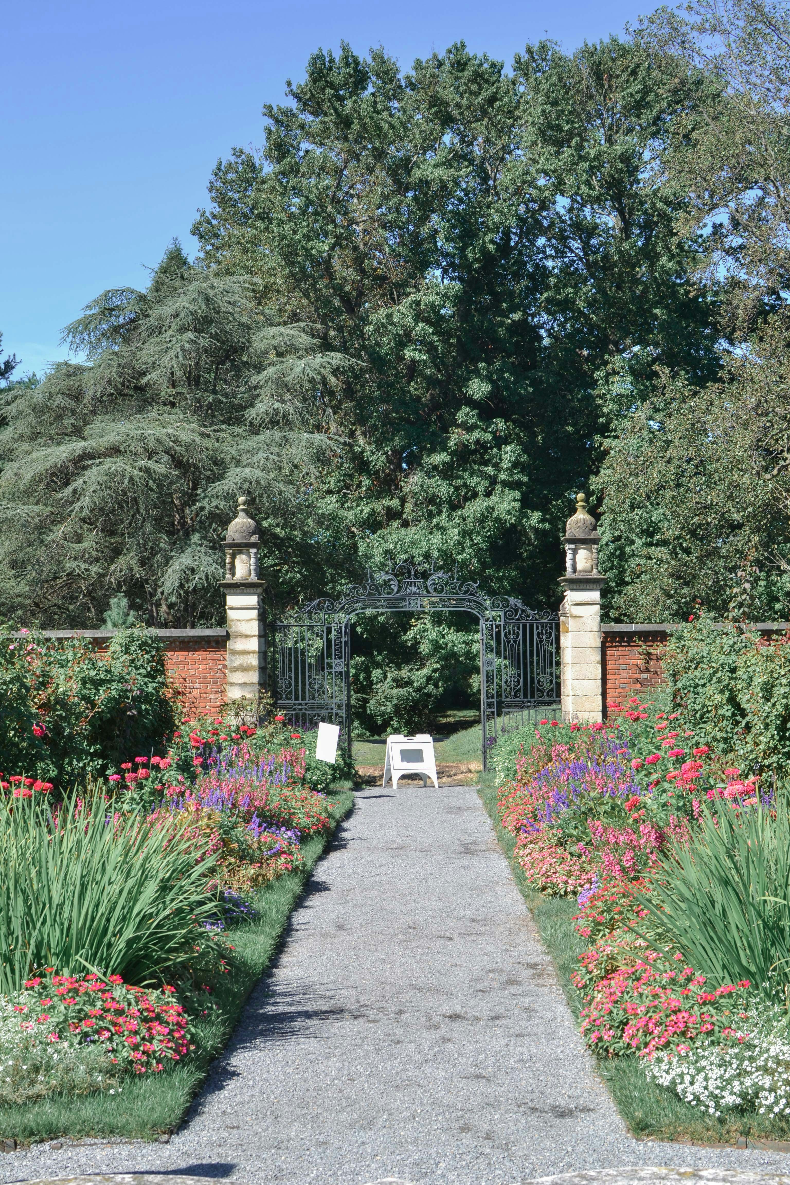A charming garden pathway leads to an ornate wrought iron gate, framed by vibrant flowers and lush greenery. The scene invites tranquility and exploration.
