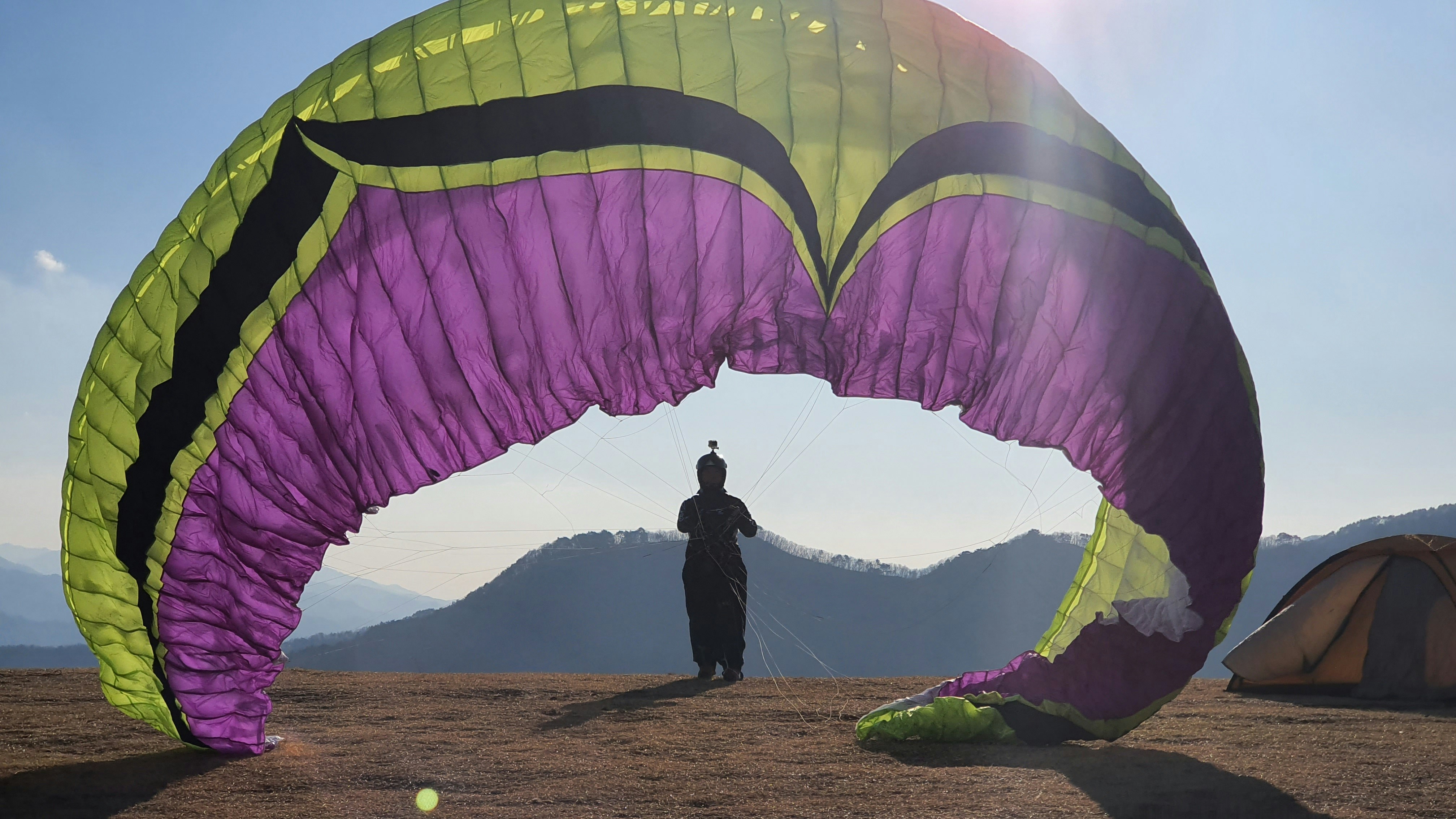 Neon canopy arch frames a solitary paraglider silhouette on a sunlit ridge