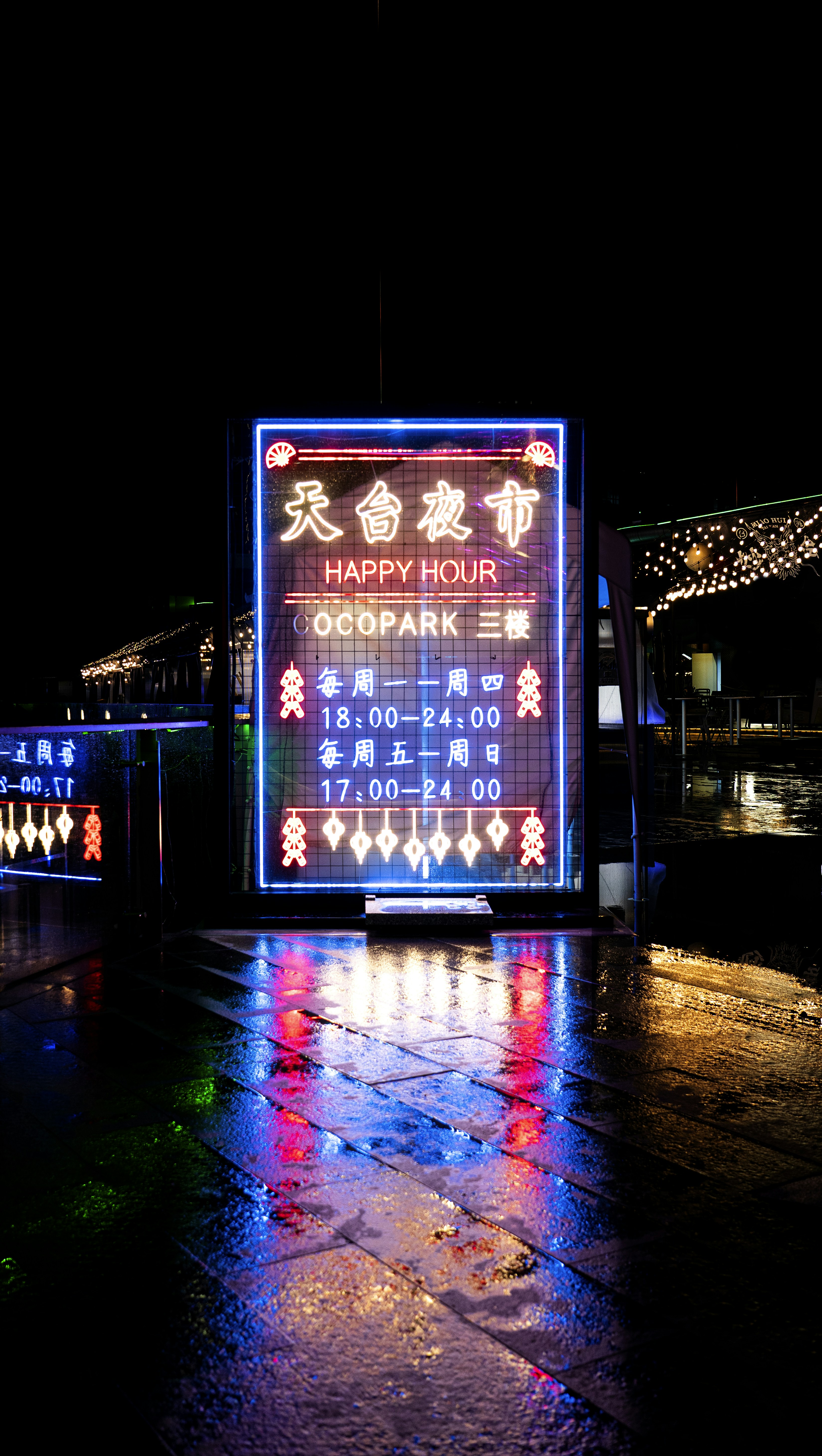 Vibrant neon sign illuminating a wet pavement at Cocopark, showcasing happy hour details. Reflections enhance the colorful ambiance of the night scene.