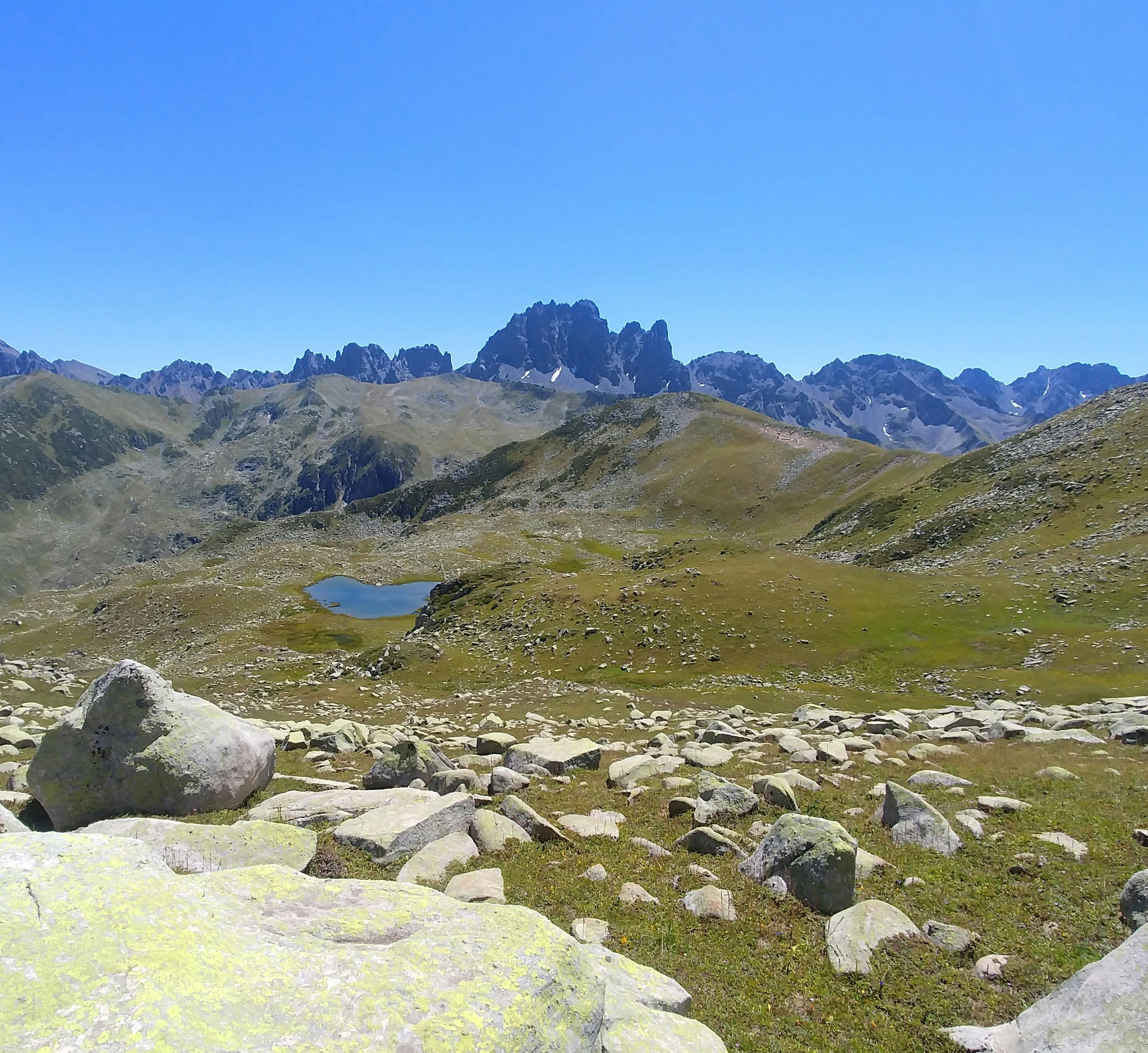 Alpine landscape featuring a small tarn bordered by rocky terrain and a jagged ridge on the horizon. The scene highlights rugged geology and an expansive blue sky.