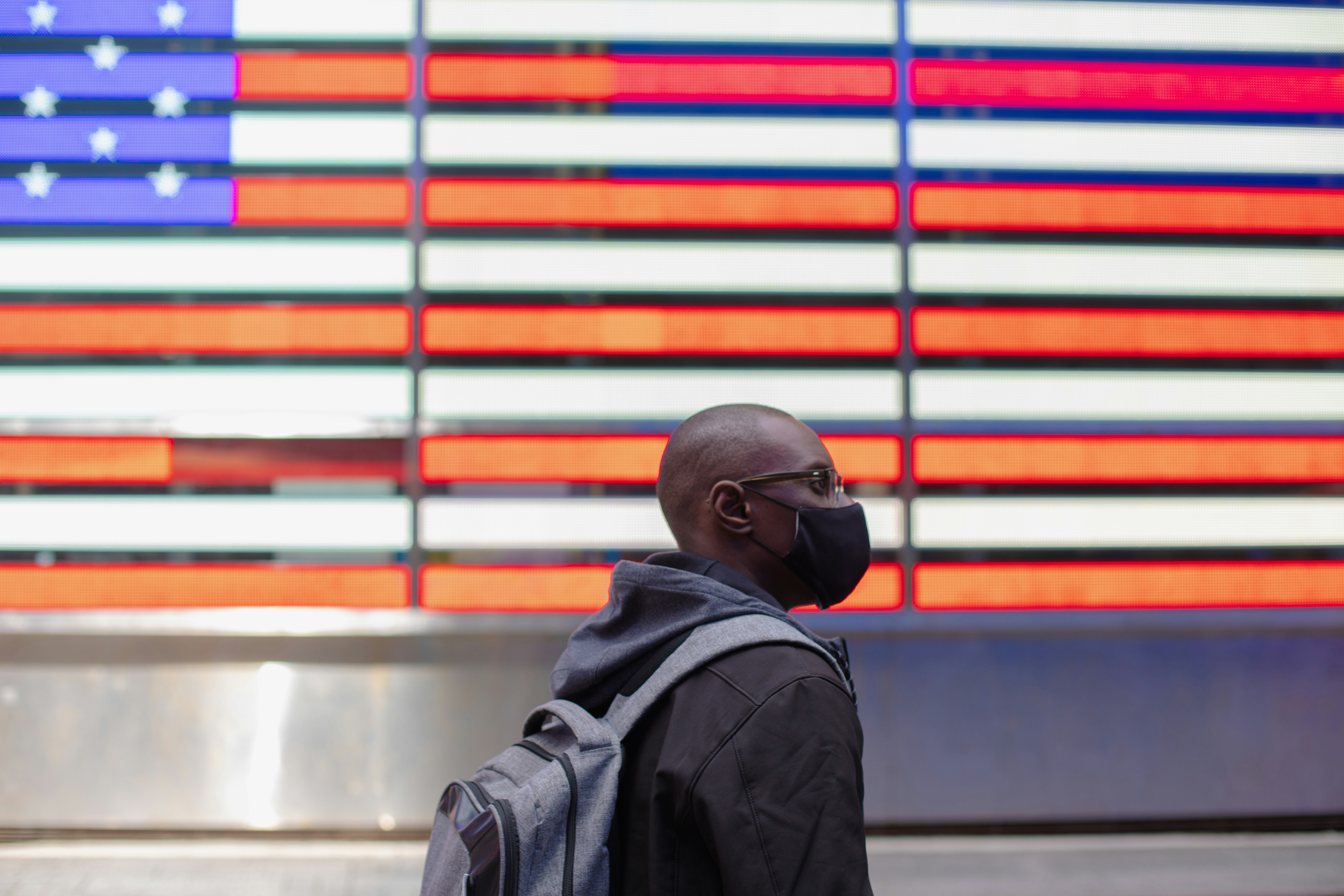 A person wearing a mask walks past a brightly lit American flag display, embodying a moment of introspection amidst urban life.