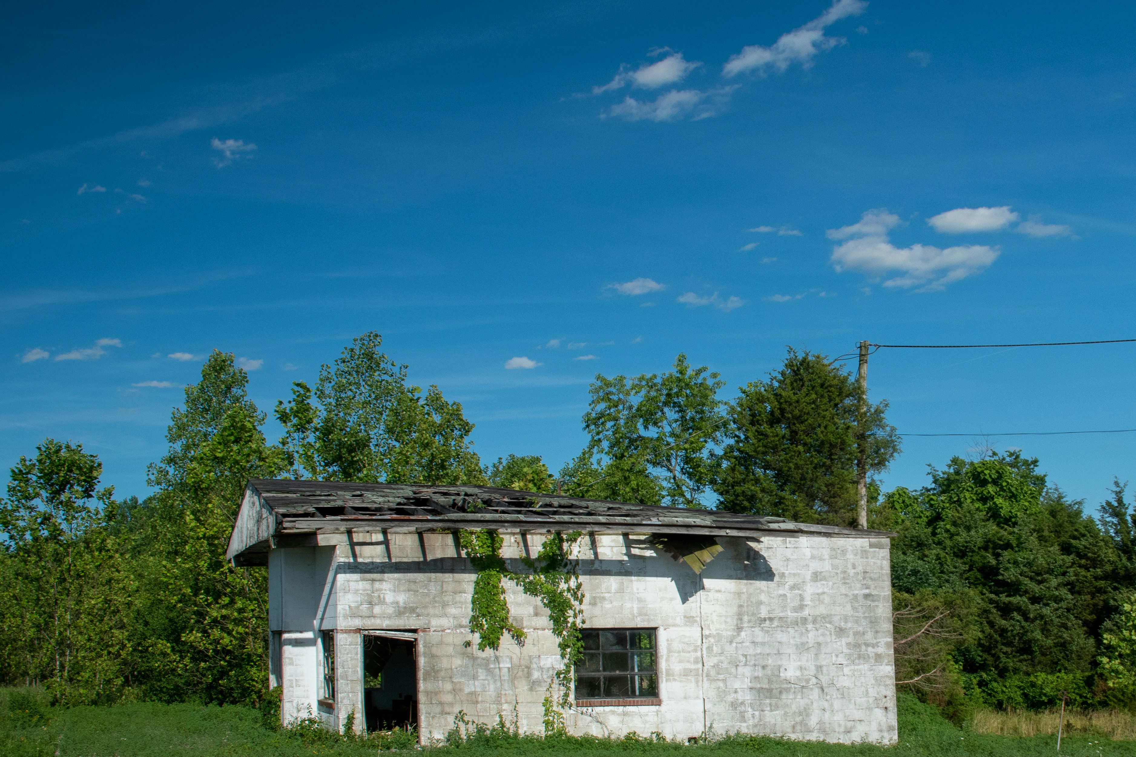Dilapidated building with a partially collapsed roof, surrounded by lush greenery under a clear blue sky.