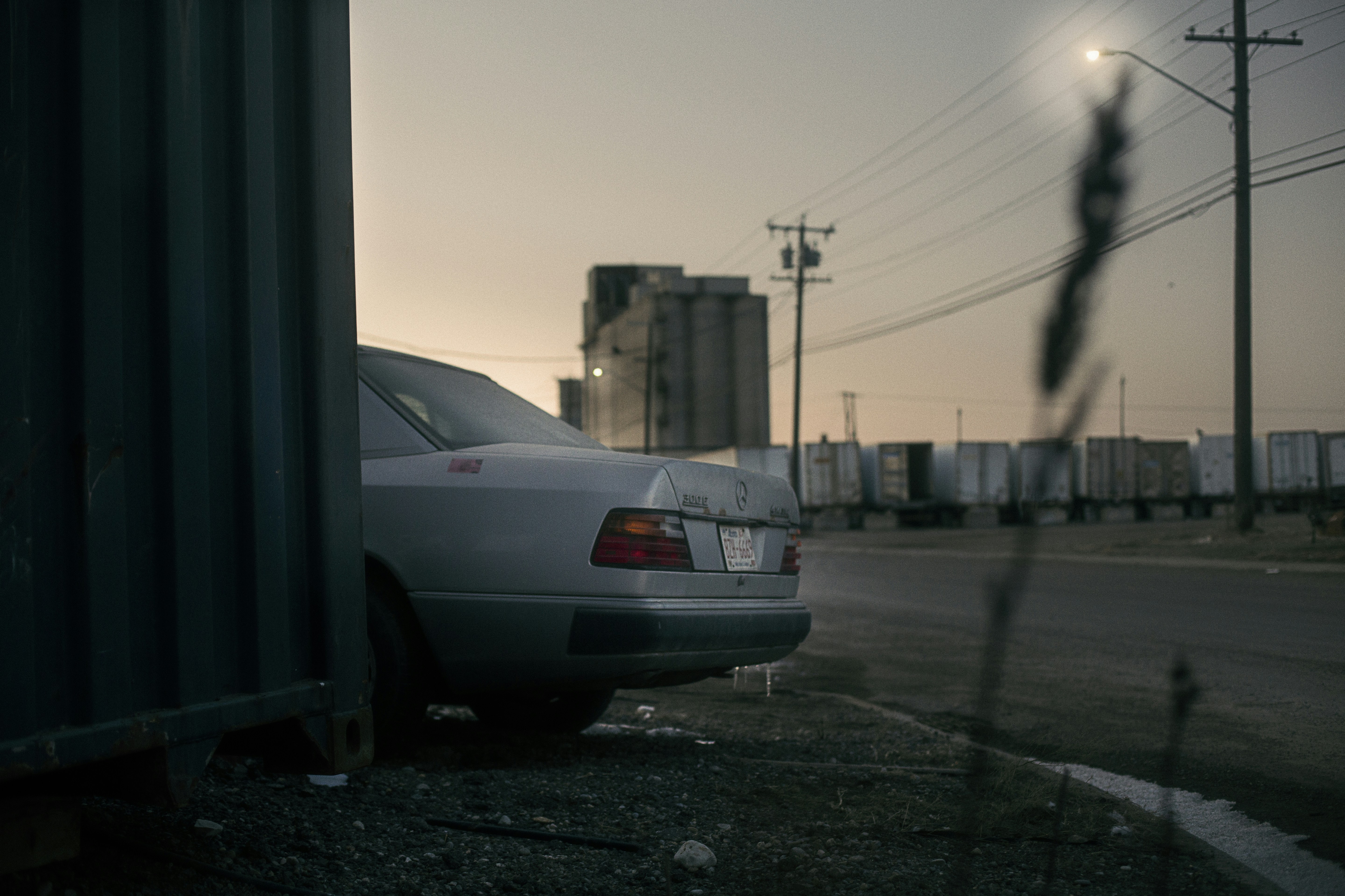 A weathered car partially obscured by a shipping container, set against a backdrop of industrial structures and power lines during twilight.