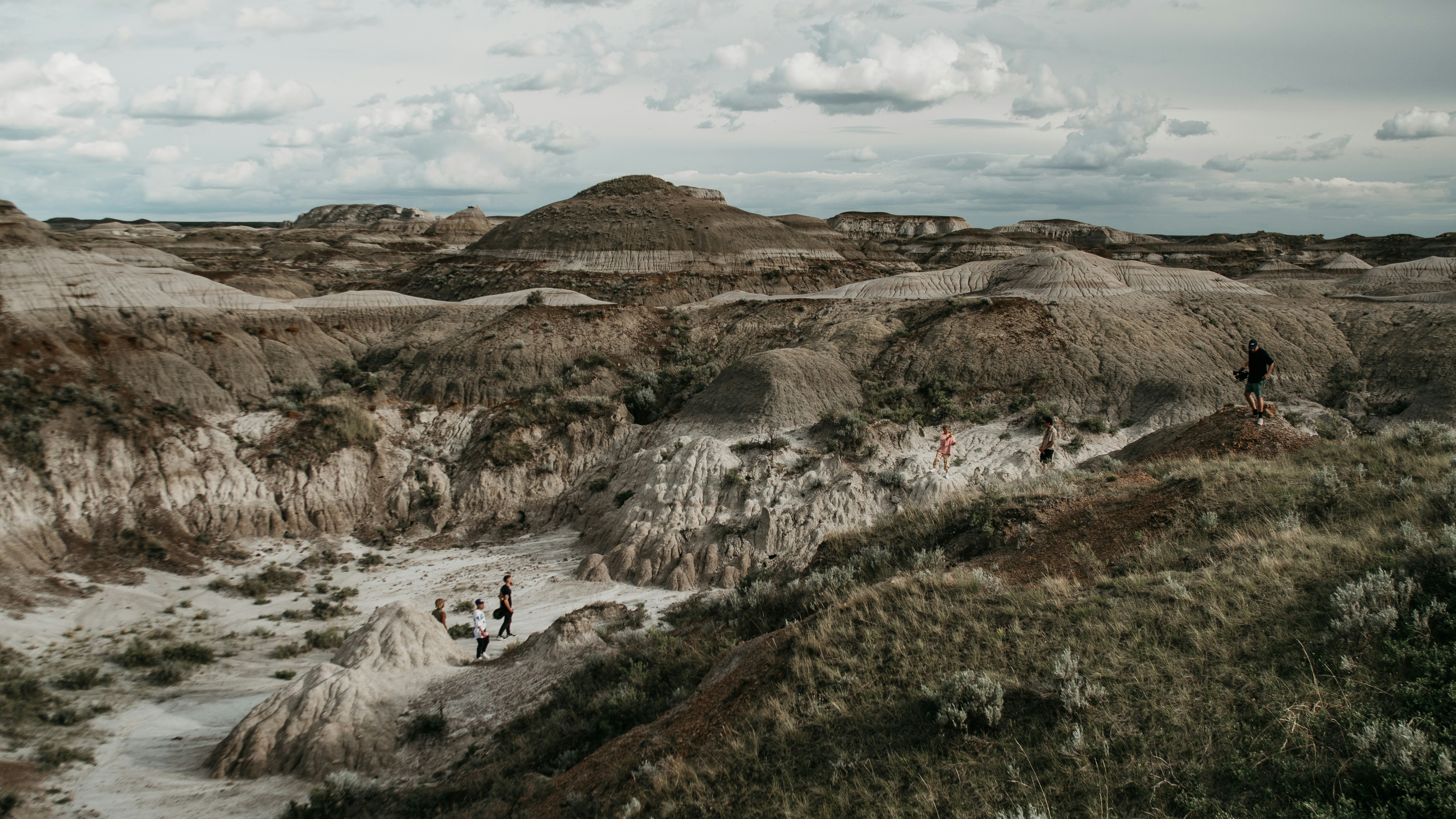 Expansive Badlands landscape with rugged terrain and hikers exploring the unique geological formations. The scene captures the interplay of light and shadow across the natural features.