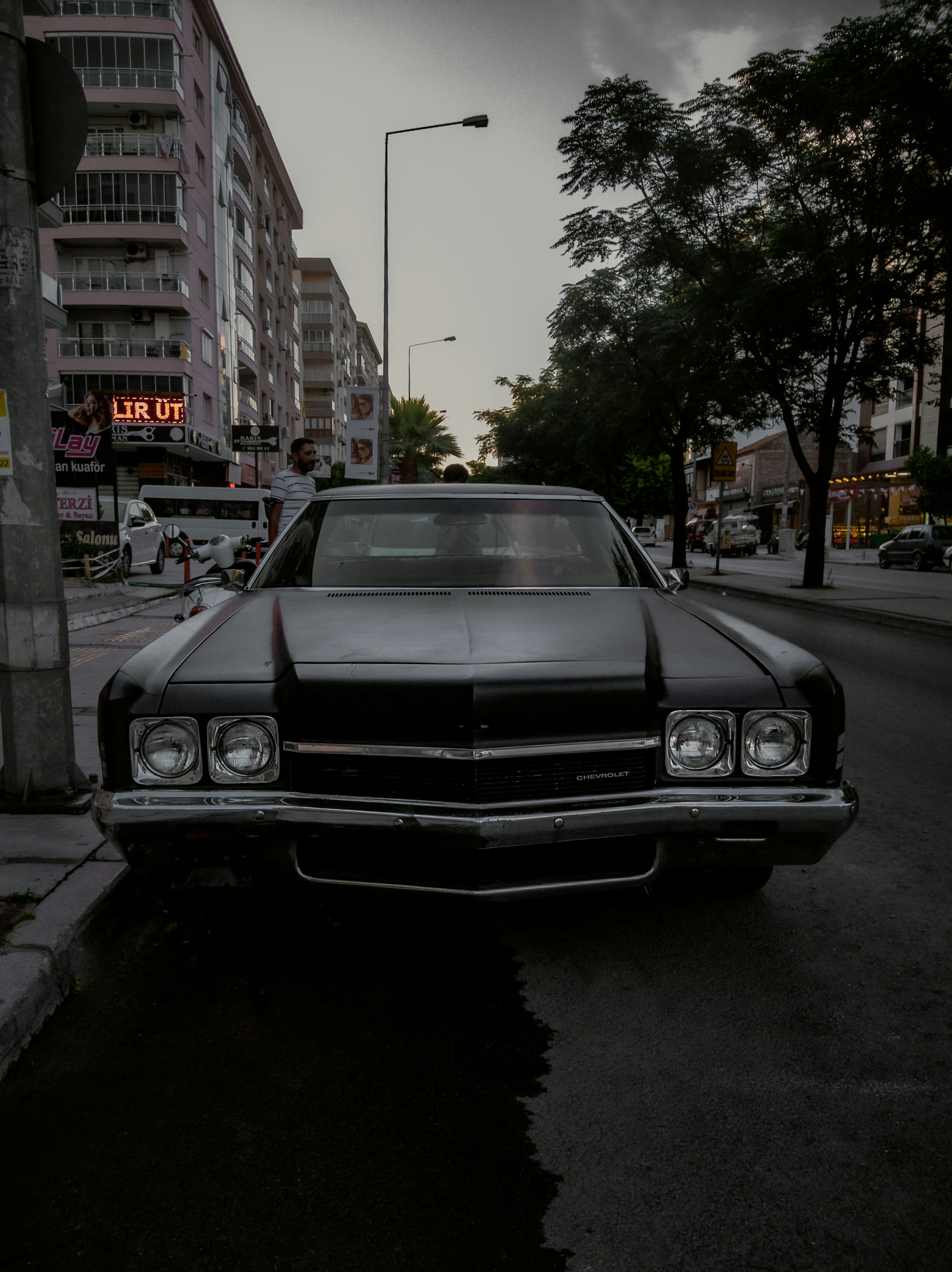 Dark vintage car dominates a dim city street at dusk, its chrome grille catching faint streetlight. Pedestrians and urban details frame the vehicle, creating a moody, cinematic moment.