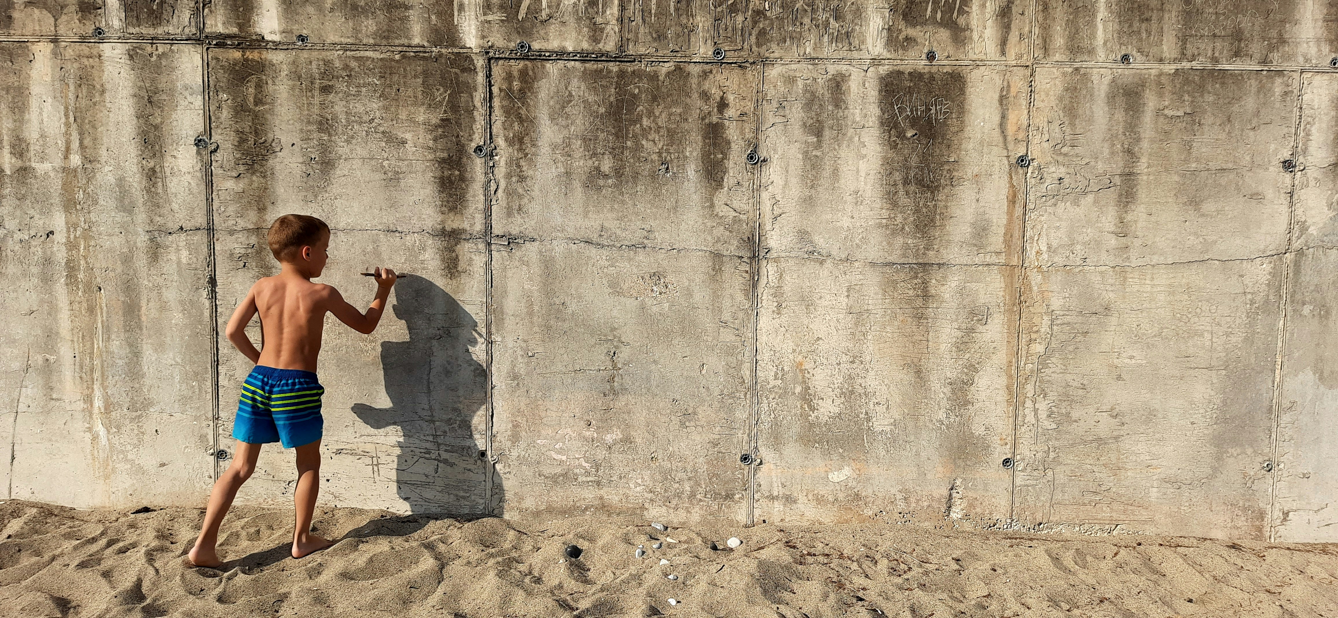 Young boy with a playful expression interacts with a textured concrete wall, casting a shadow on the sandy ground.