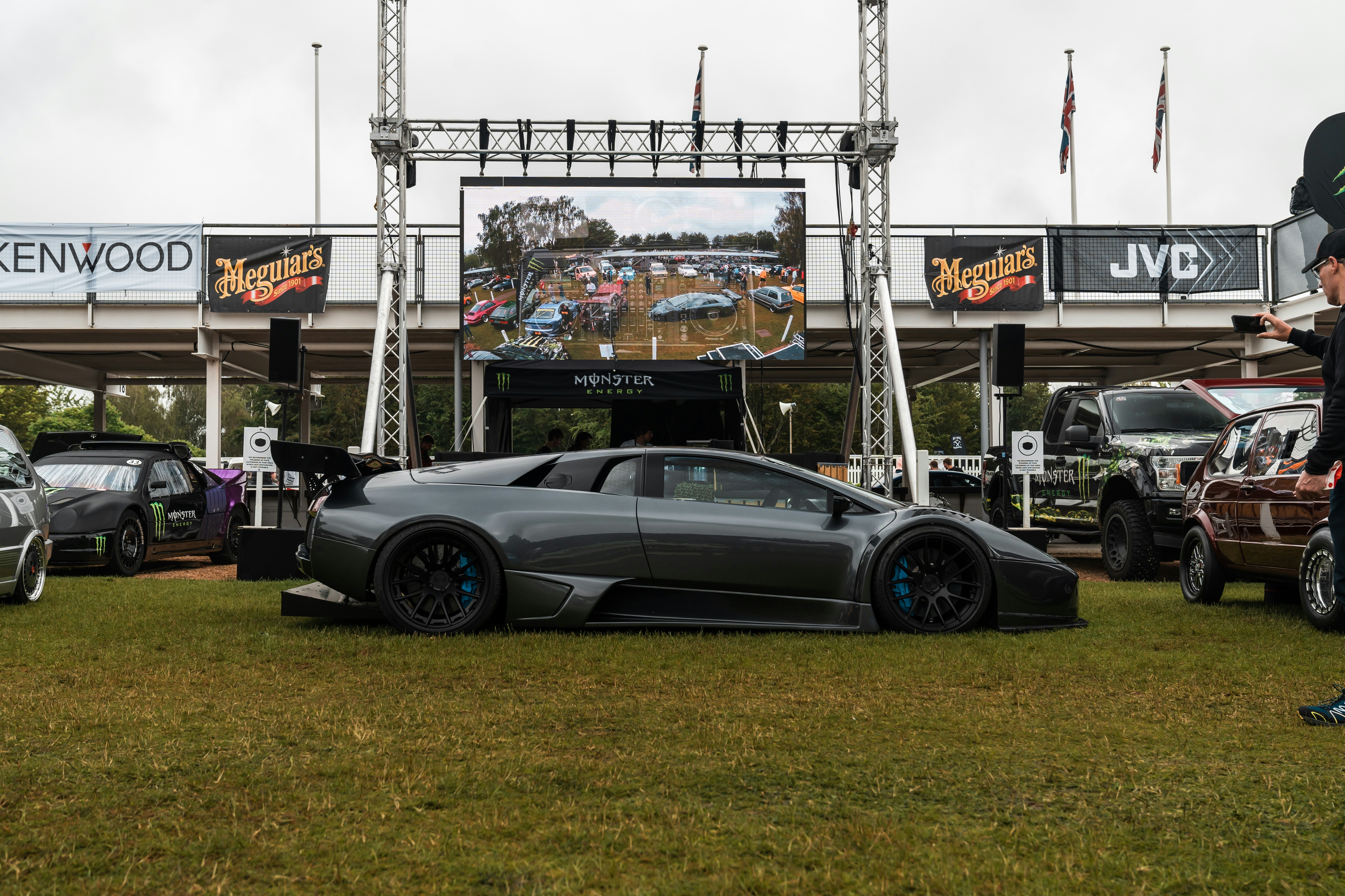Sleek gray Lamborghini positioned prominently in an automotive exhibition, surrounded by modified vehicles and a large screen displaying event highlights.