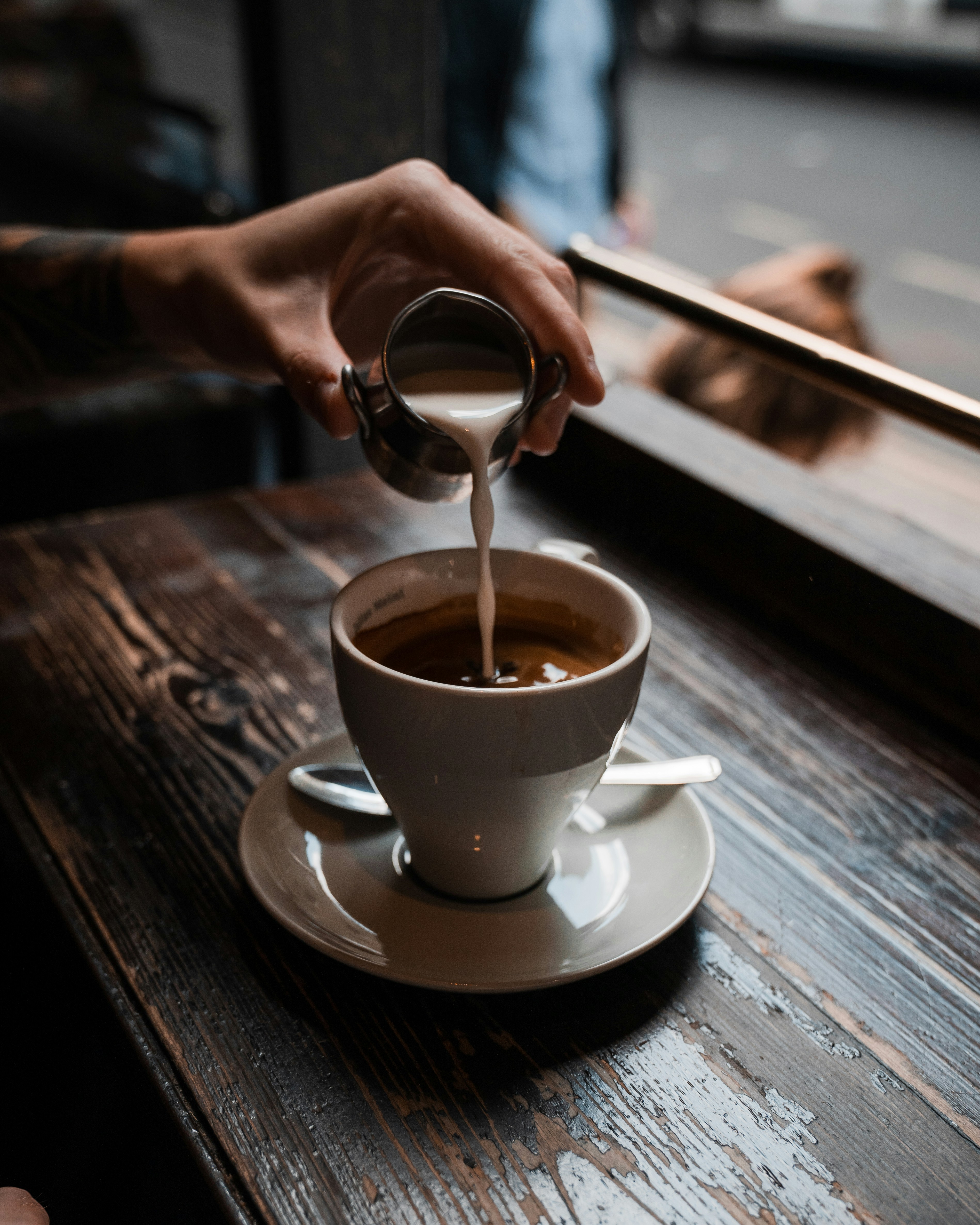 A hand pours milk into a steaming cup of coffee, capturing the intimate moment of coffee preparation in a cozy café setting.