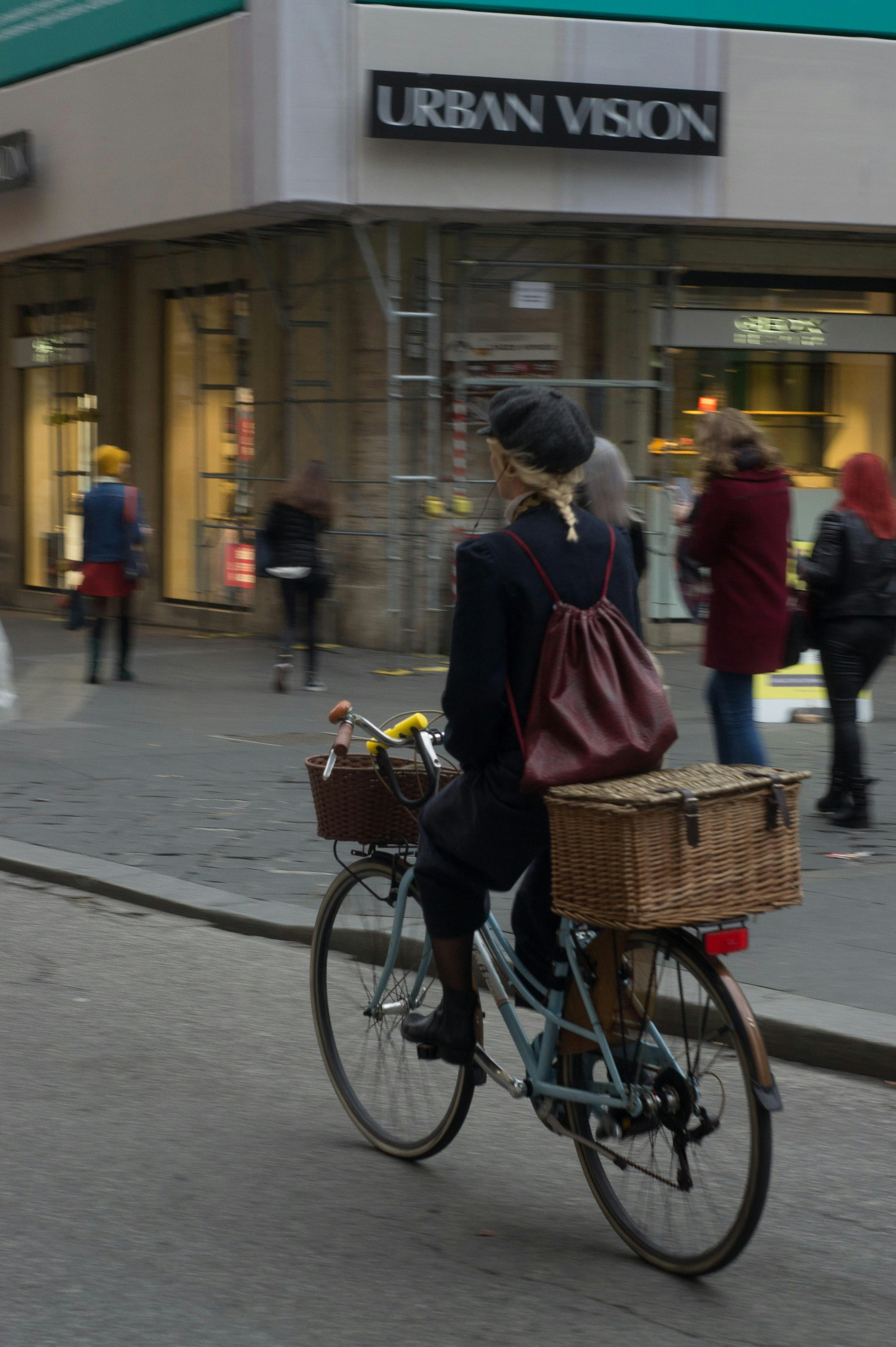 Cyclist with wicker baskets on a blue bicycle rides along a city street, while pedestrians pass by and an Urban Vision storefront looms in the background.