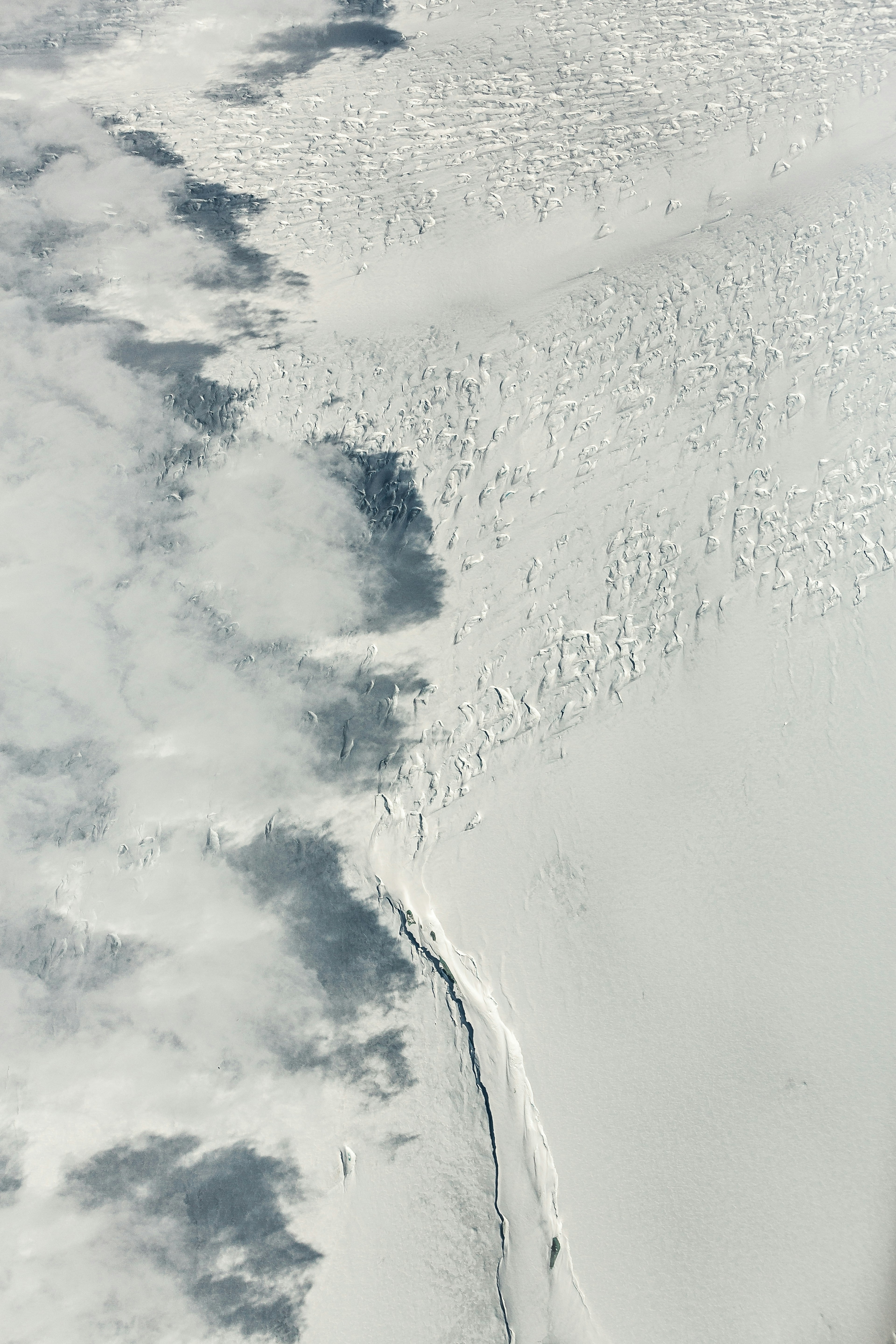 Aerial view of a vast icy landscape, showcasing intricate patterns of snow and ice juxtaposed with soft clouds. The scene evokes a sense of serene isolation.
