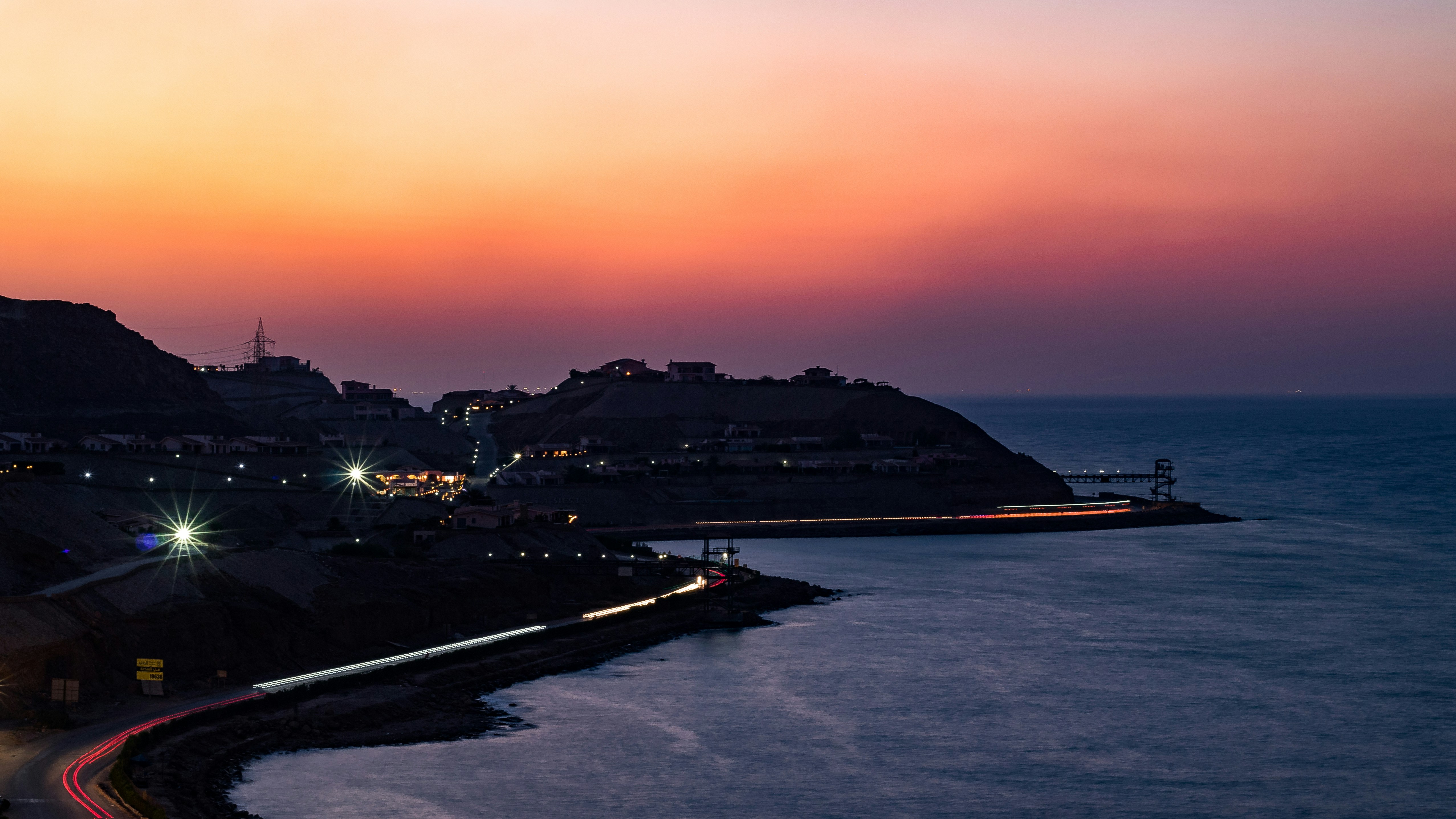 A serene coastal landscape at twilight, showcasing a winding road with light trails and a calm sea under a gradient sky. 