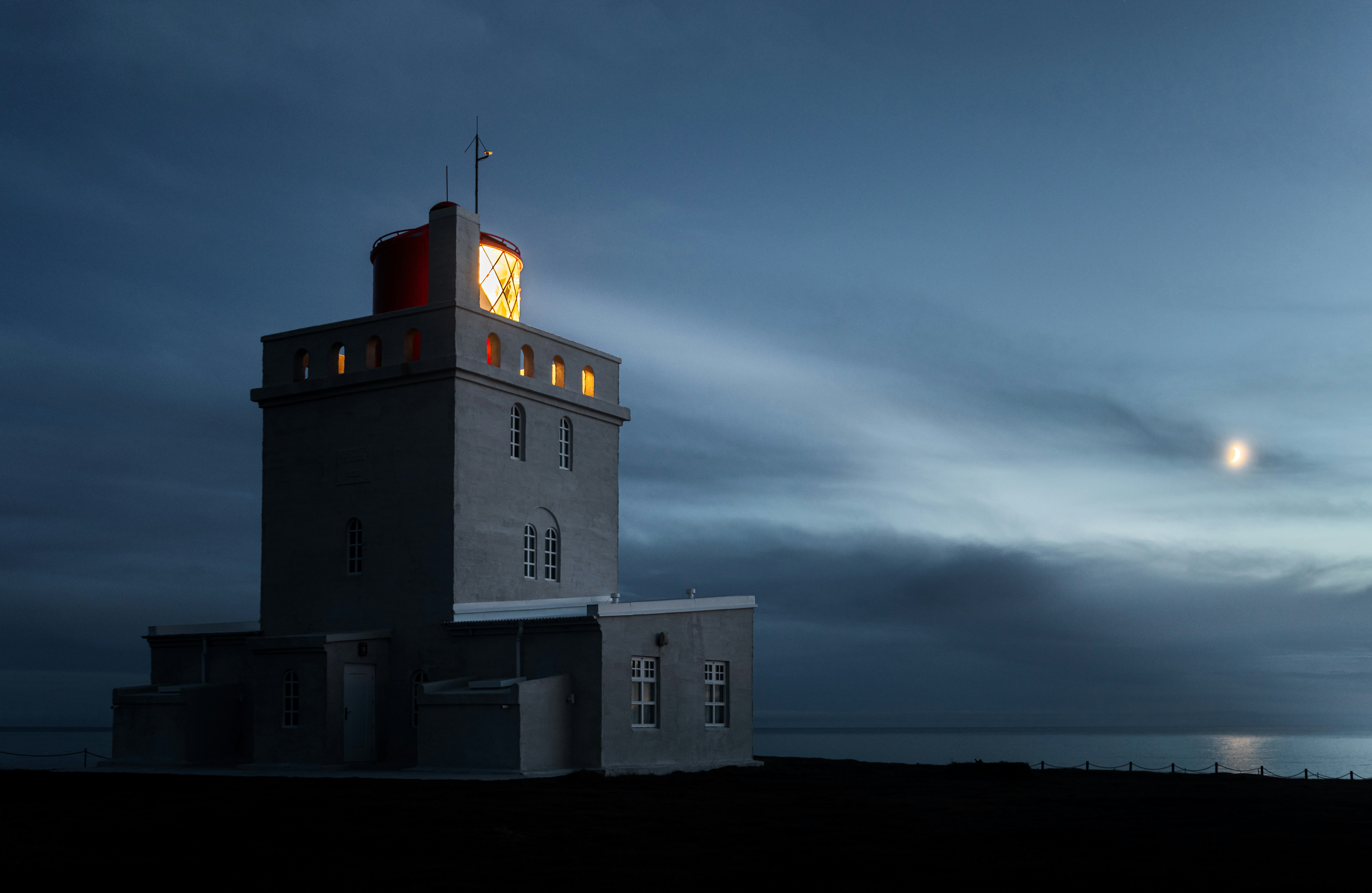 A solitary lighthouse stands against a twilight sky, its lantern glowing warmly amidst the cool hues of dusk. The calm ocean reflects the fading light.