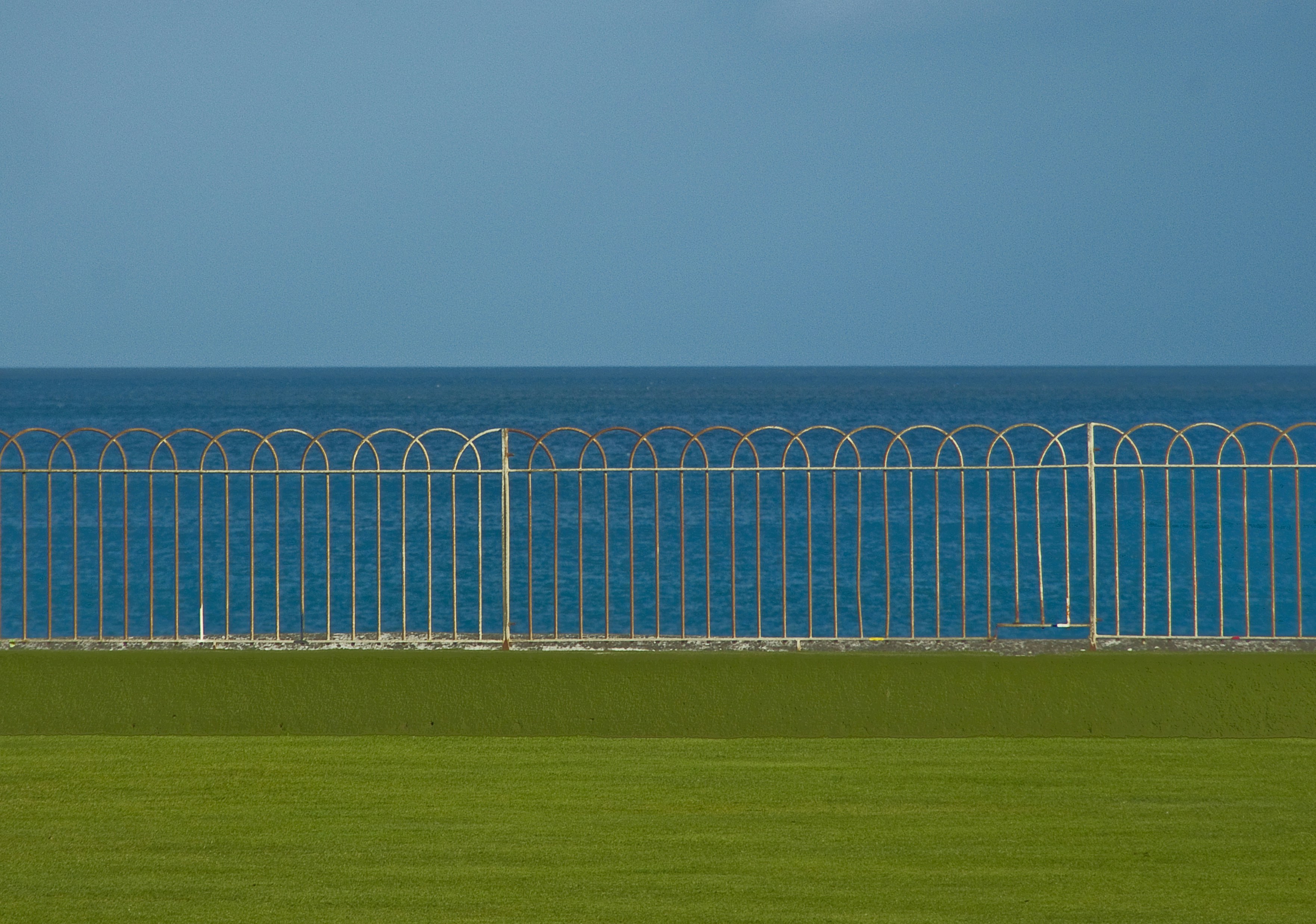 Lime-green lawn beside an arched metal fence, with a calm blue sea on the horizon.