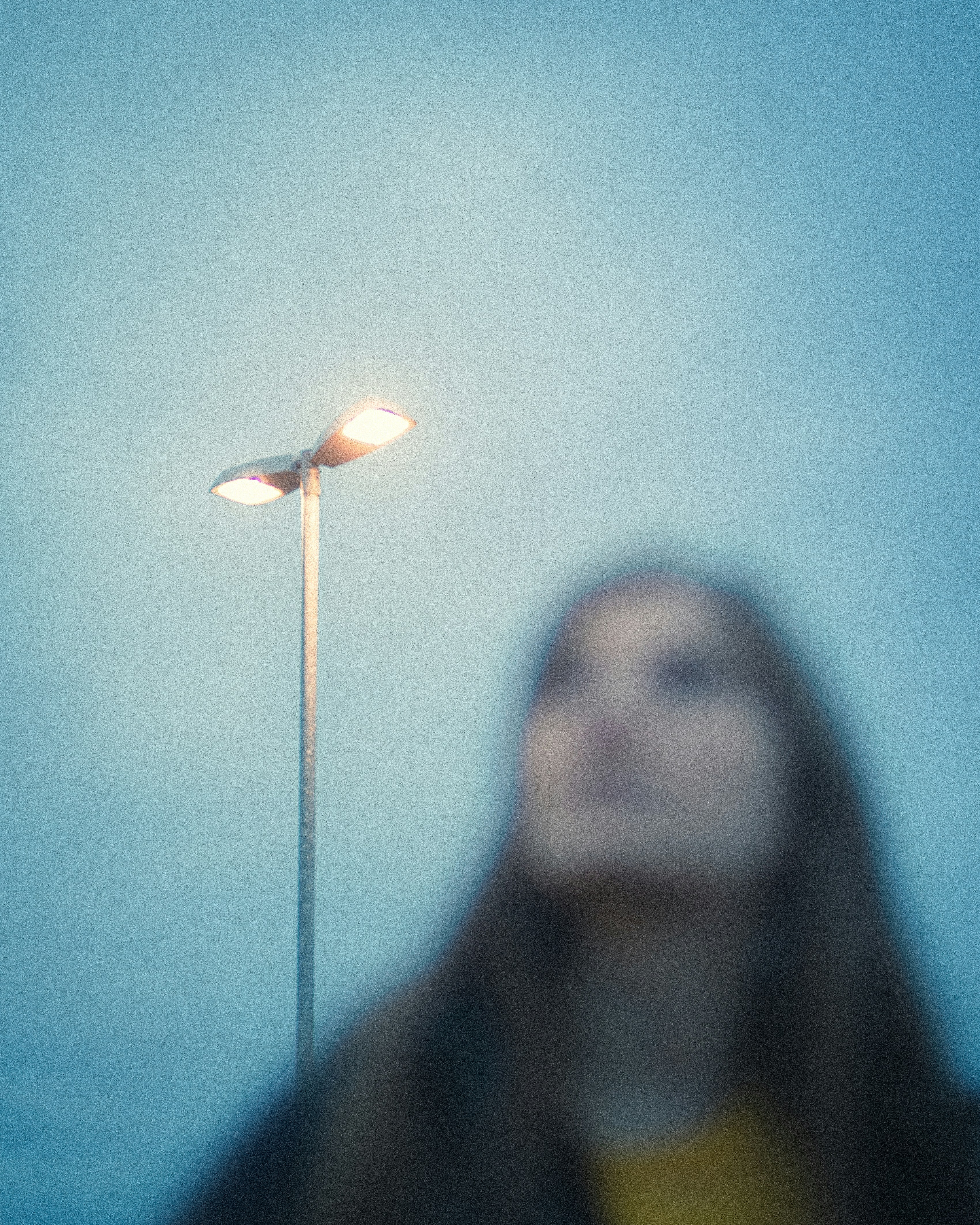 A blurred figure stands beneath a streetlamp, creating a moody atmosphere against a twilight sky.