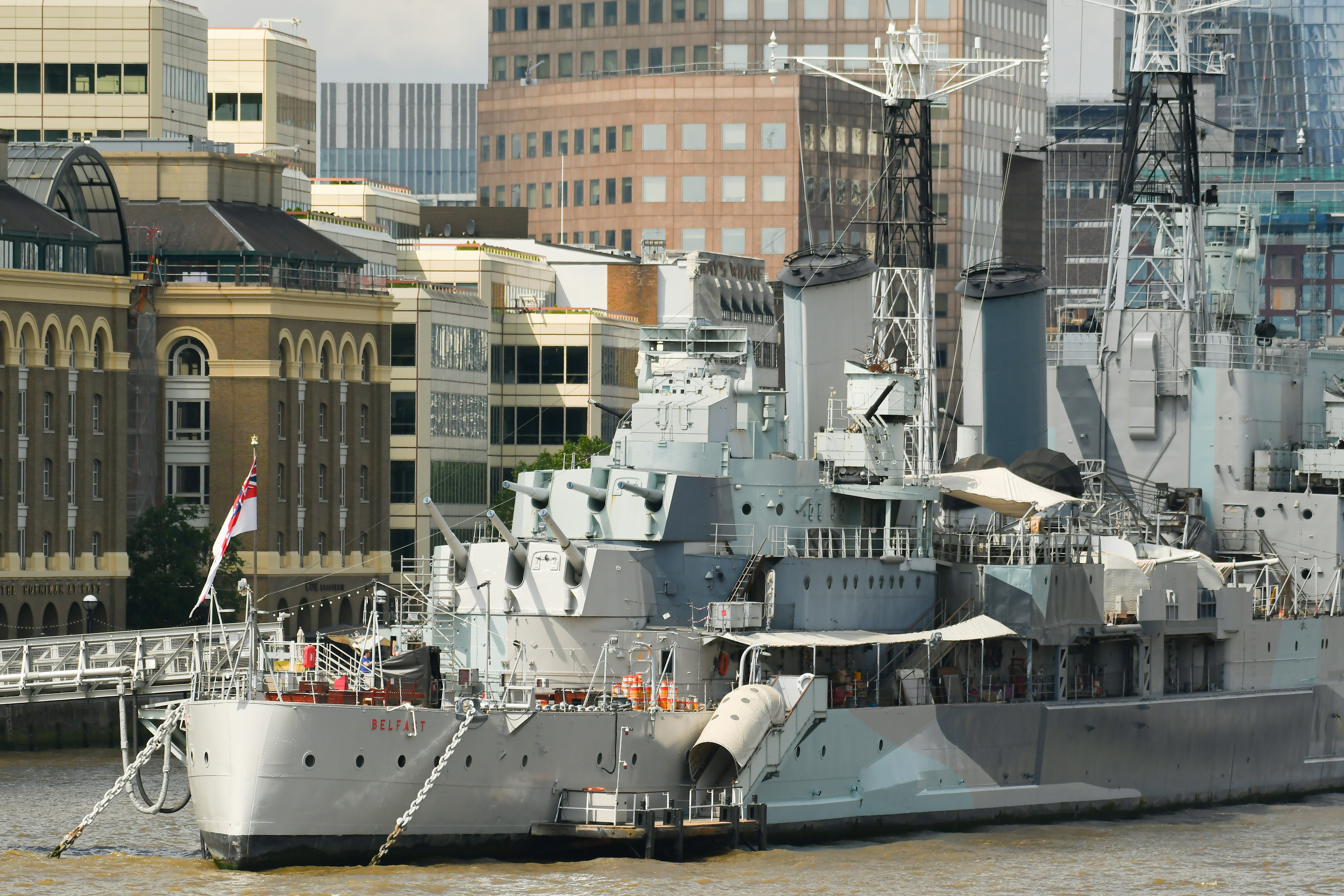 Historic naval ship docked beside modern buildings along the river, showcasing a blend of maritime history and contemporary architecture.