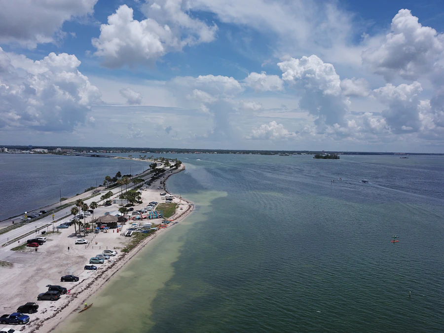 Aerial view of a narrow causeway lined with parked cars and palm trees, stretching into a broad bay under towering cumulus clouds. Shallow green water hugs the shoreline beside deeper blue water.